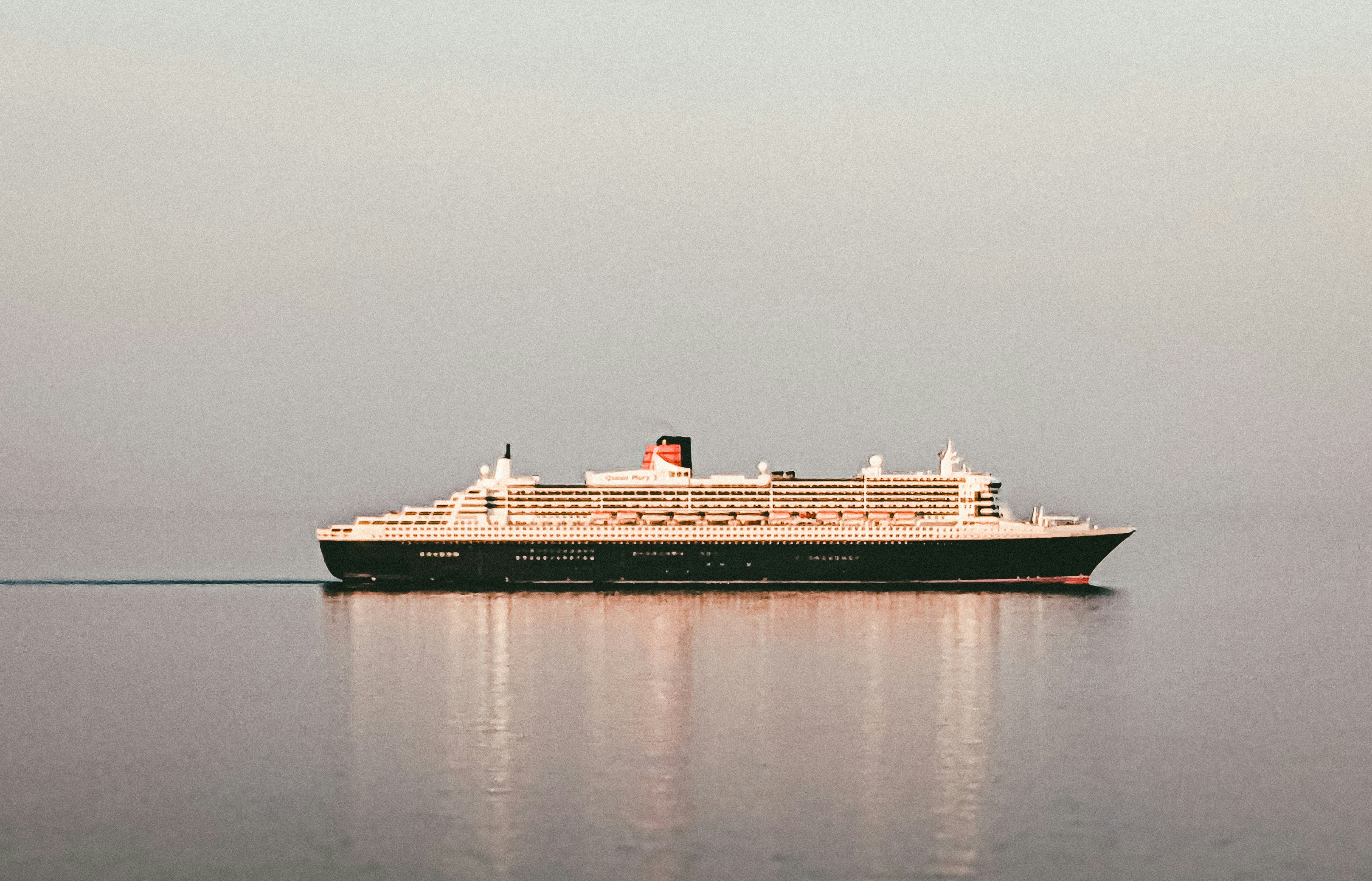 A solitary cruise ship glides on glassy water, its reflection stretching beneath a pale horizon; the scene emphasizes symmetry and quiet maritime motion. This is a photographic capture of a vessel on smooth seas.