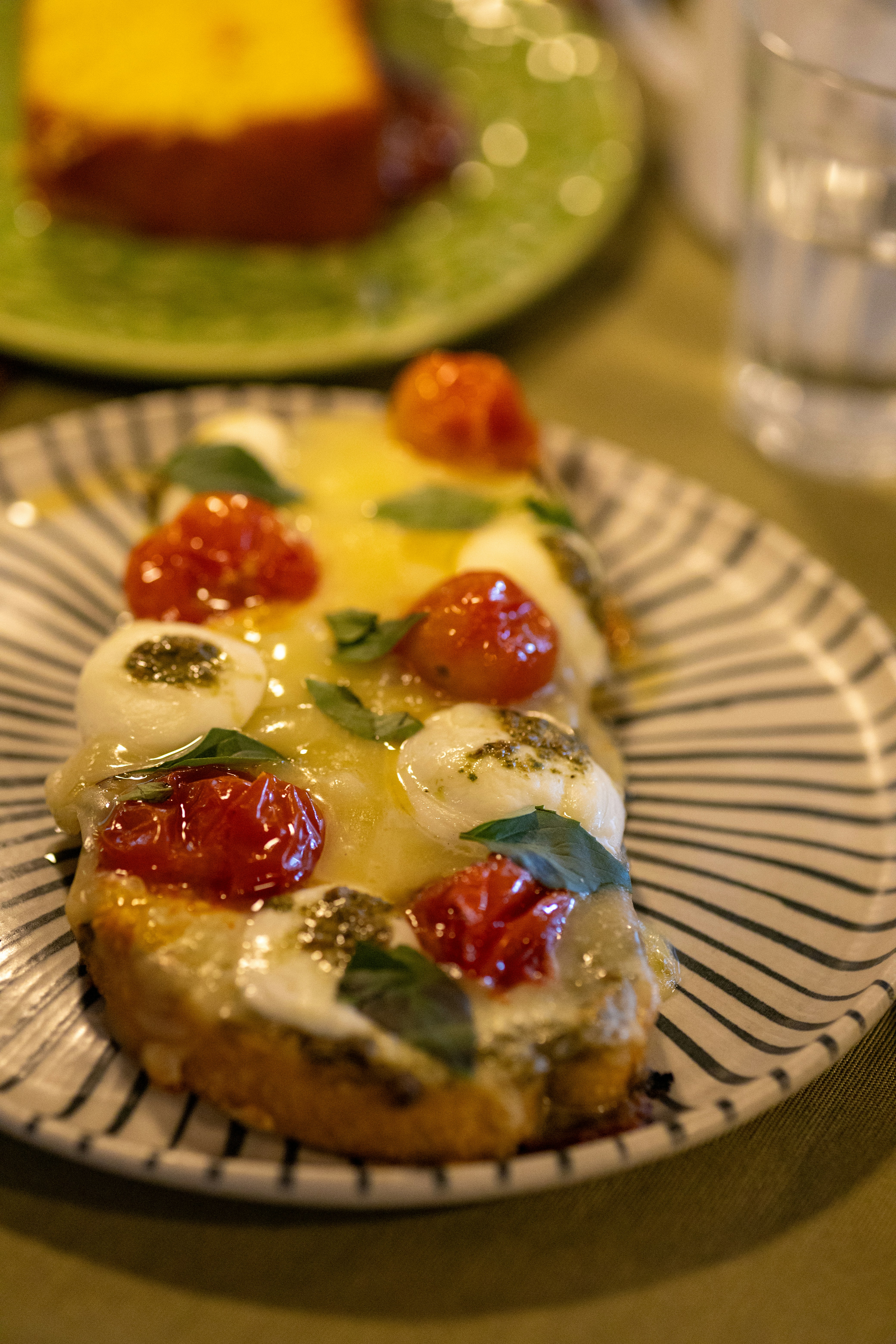 A close-up of a topped slice on a striped plate, with melted cheese, cherry tomatoes, and fresh basil, accompanied by a glass of water in soft focus.