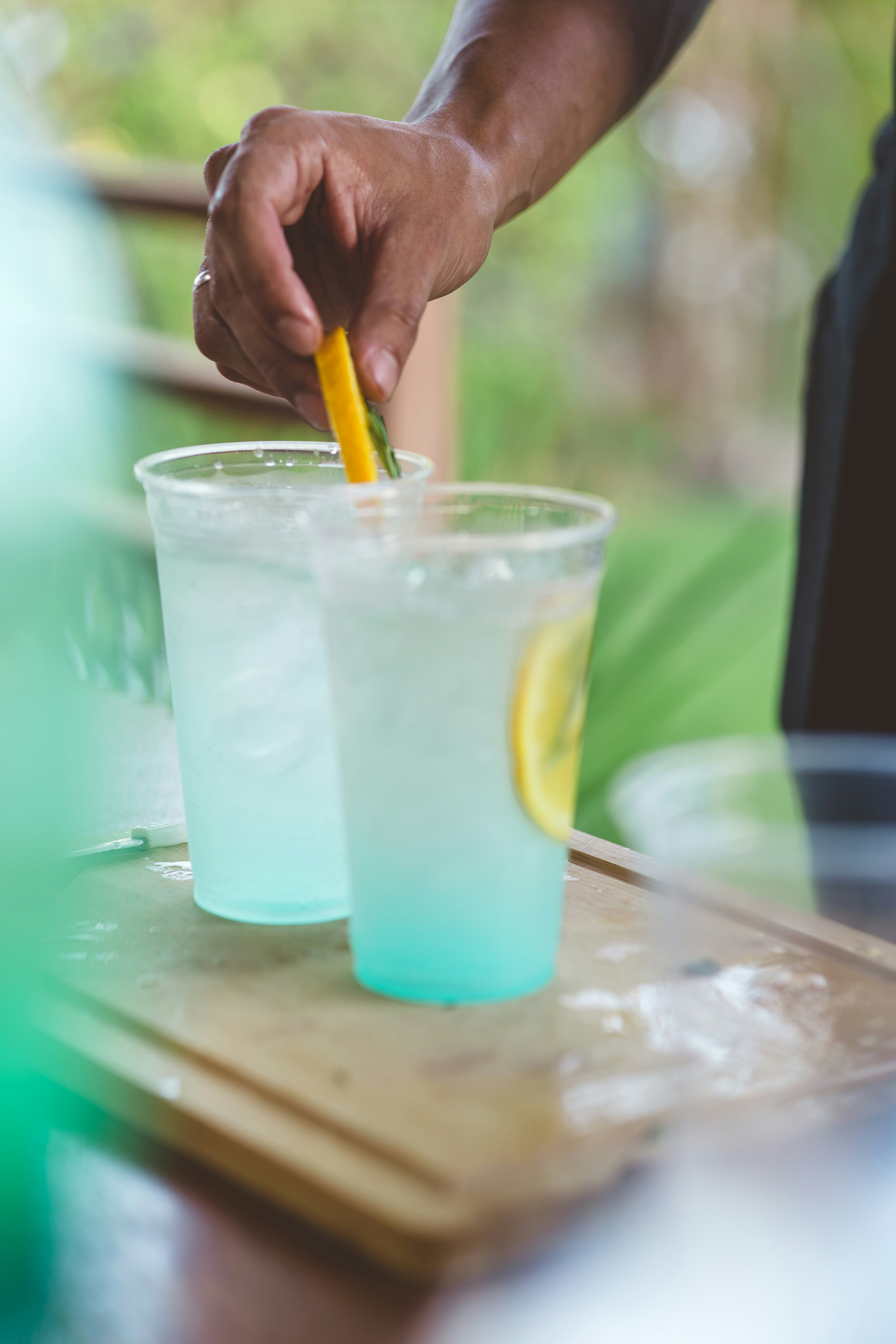 Hand drizzles a citrus glow: blue-tinted drinks on a wooden tray