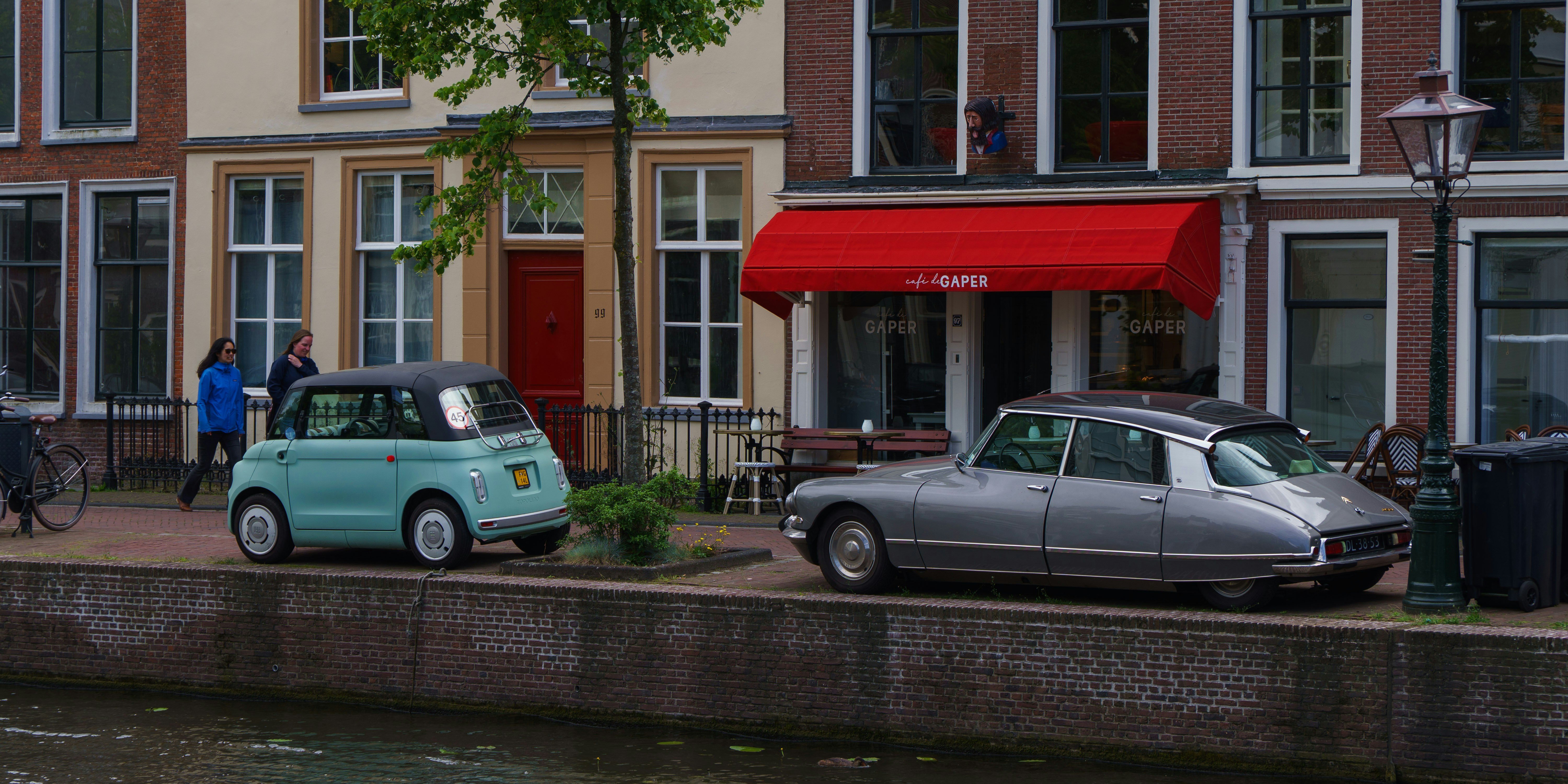Pastel teal microbus and a vintage coupe parked along a canal with brick buildings and a red awning storefront in the background.