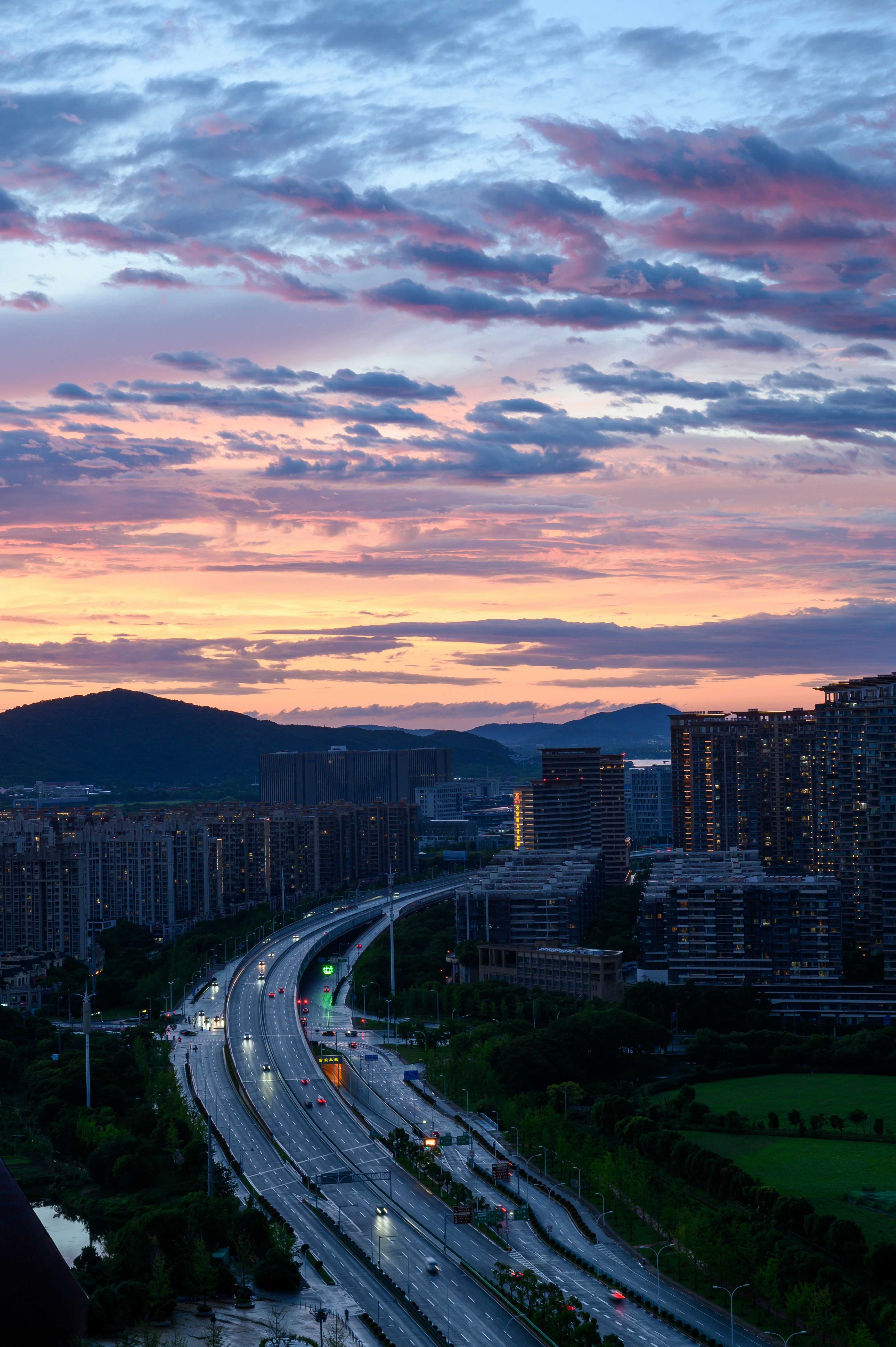 City skyline with a curving highway beneath a pastel sunset, highlighting the elevated roadway and distant hills; appears as a photographic capture of urban life at dusk.