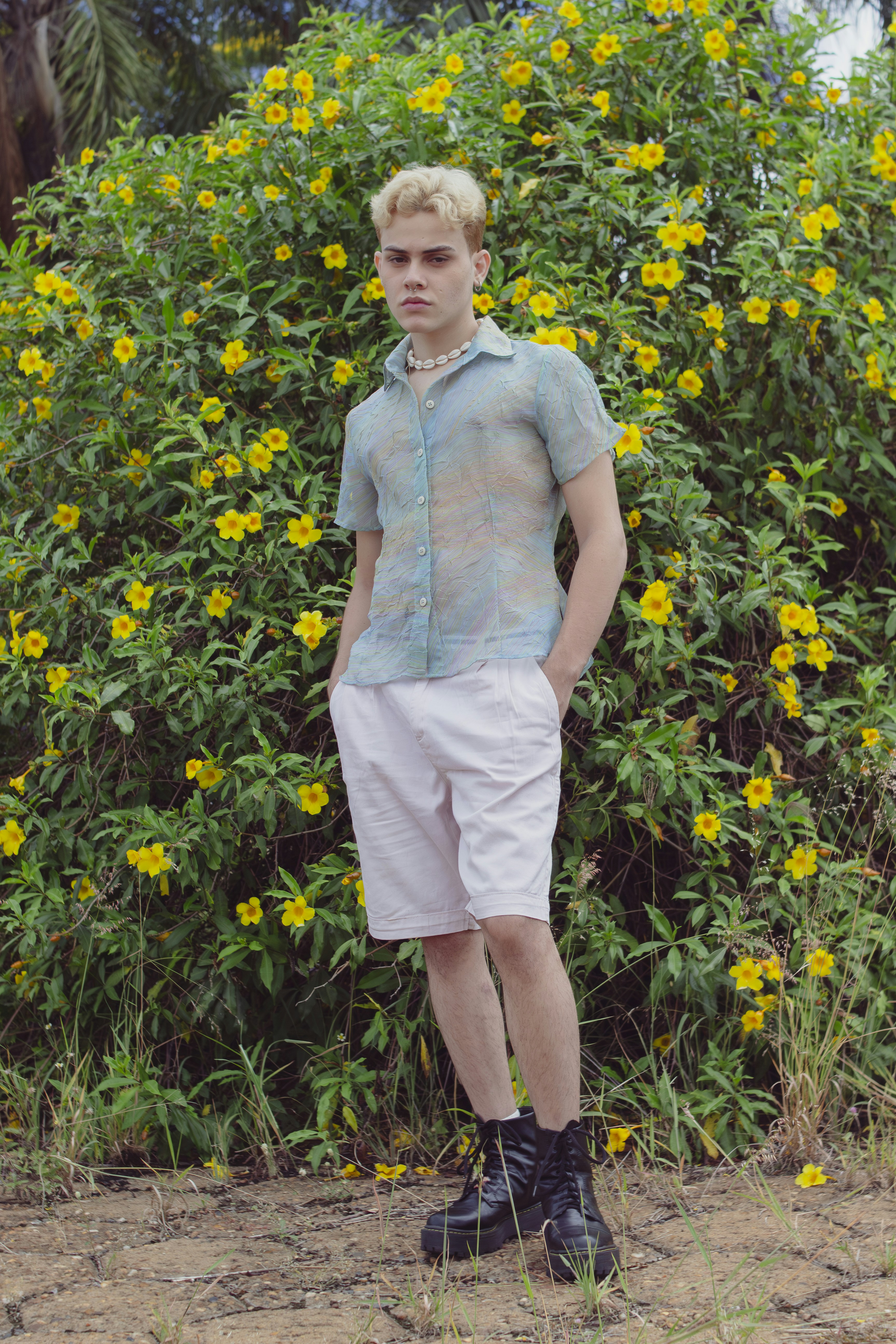 Young man stands framed by lush yellow flowers, wearing a light blue shirt, white shorts, and black boots; commands the frame with a relaxed stance in a sunlit garden. This is a candid portrait featuring casual summer style.