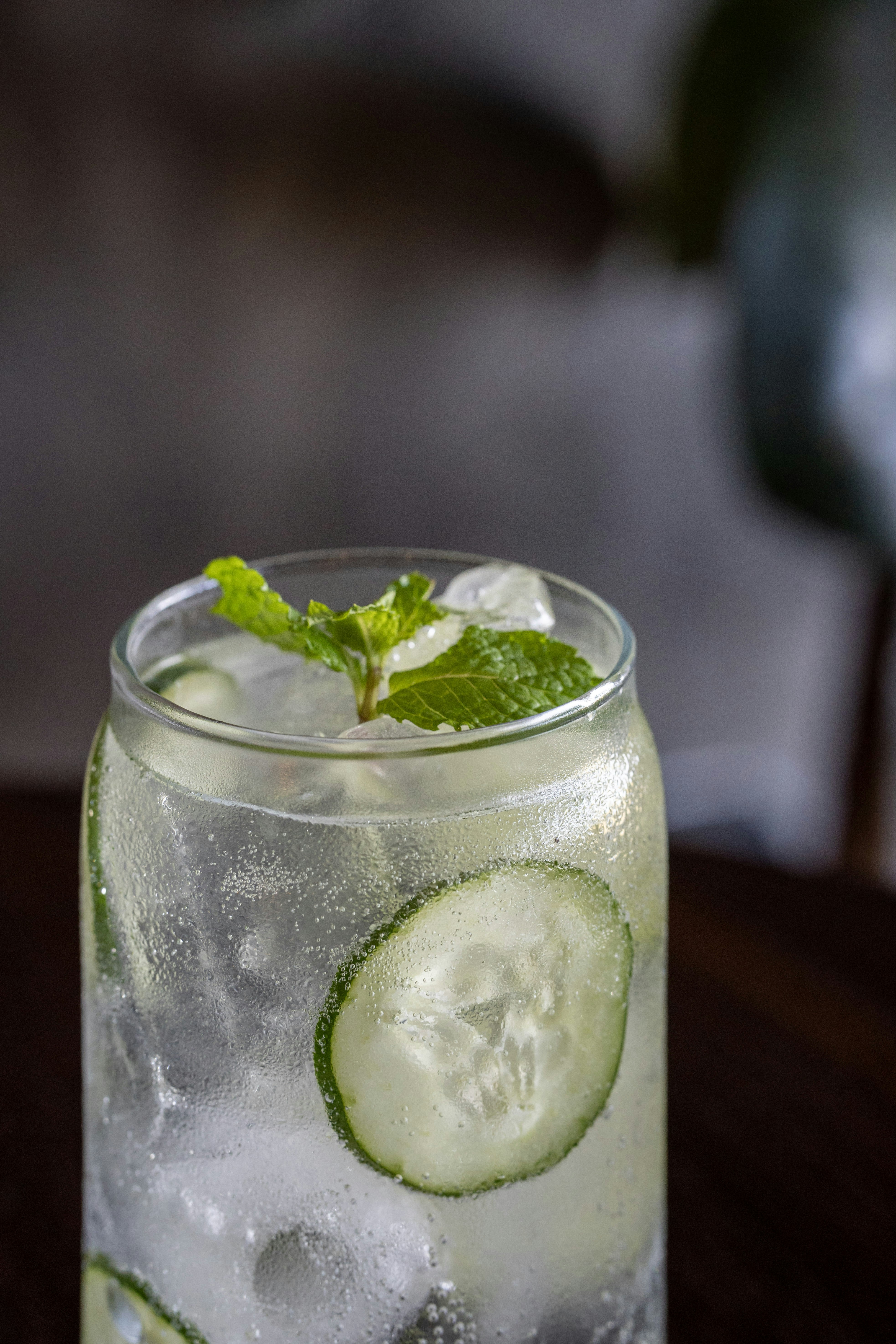 A chilled cucumber-lime beverage with mint garnish sits in a clear glass, ice cubes and condensation visible; the scene emphasizes refreshment and beverage presentation in soft studio lighting. This is a photography shot of a drink (not a logo or illustration).