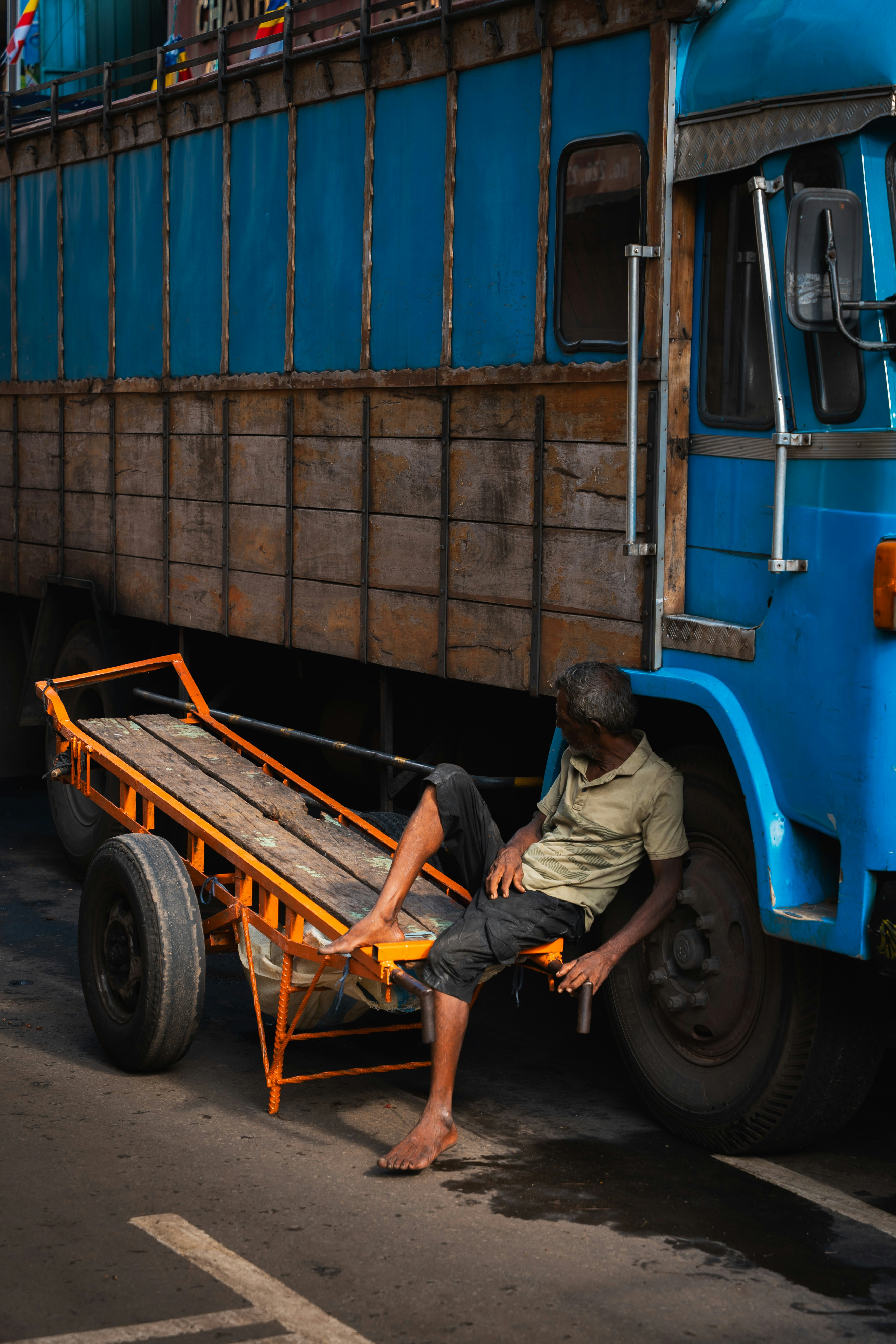 Man resting on a wheeled orange cart beside a blue cargo truck