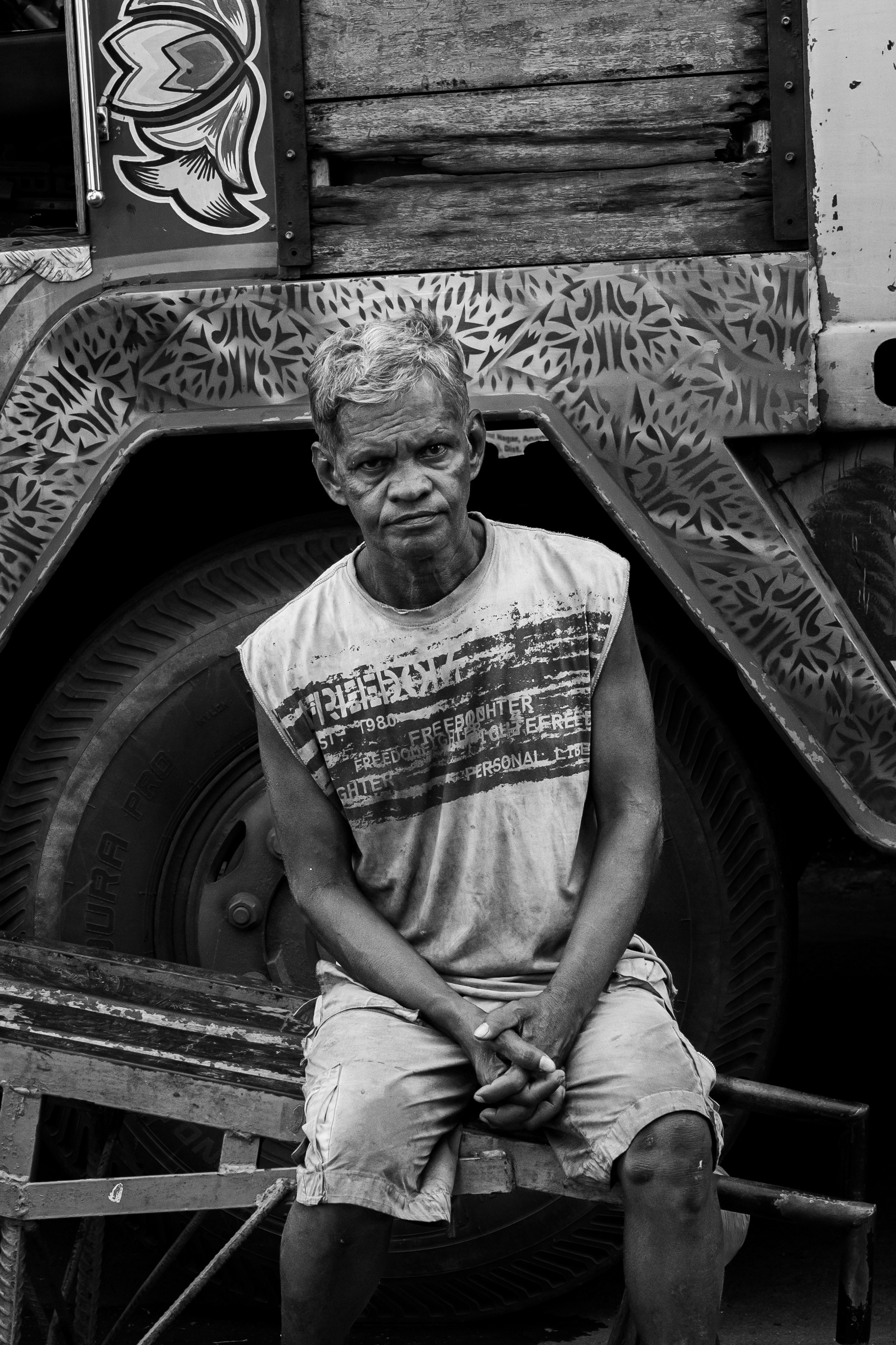 Man in a weathered sleeveless shirt sits before a graffiti-painted truck wheel