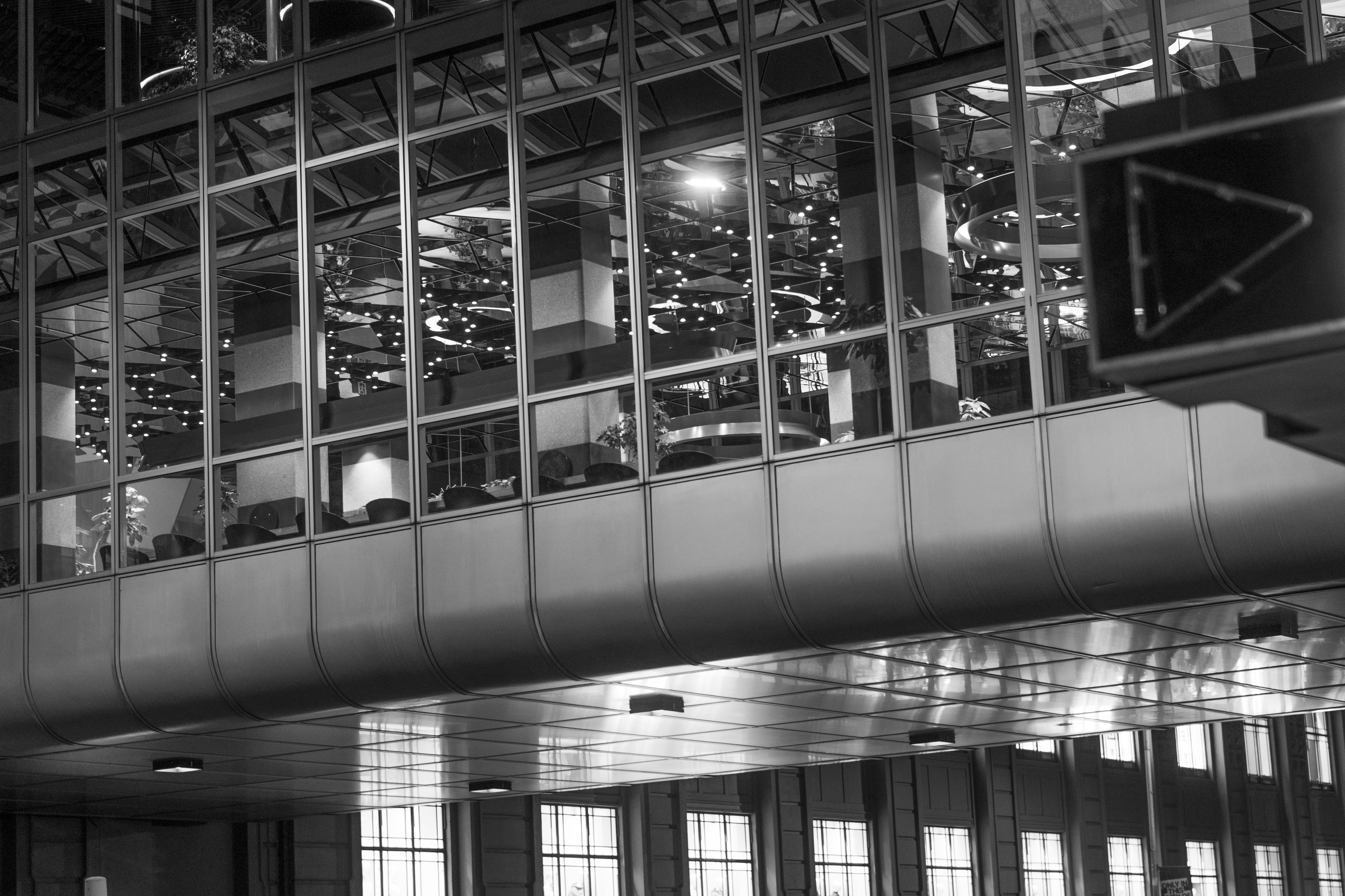A grayscale urban facade with intersecting glass panels and architectural lines, capturing reflections and ceiling details of a modern building.