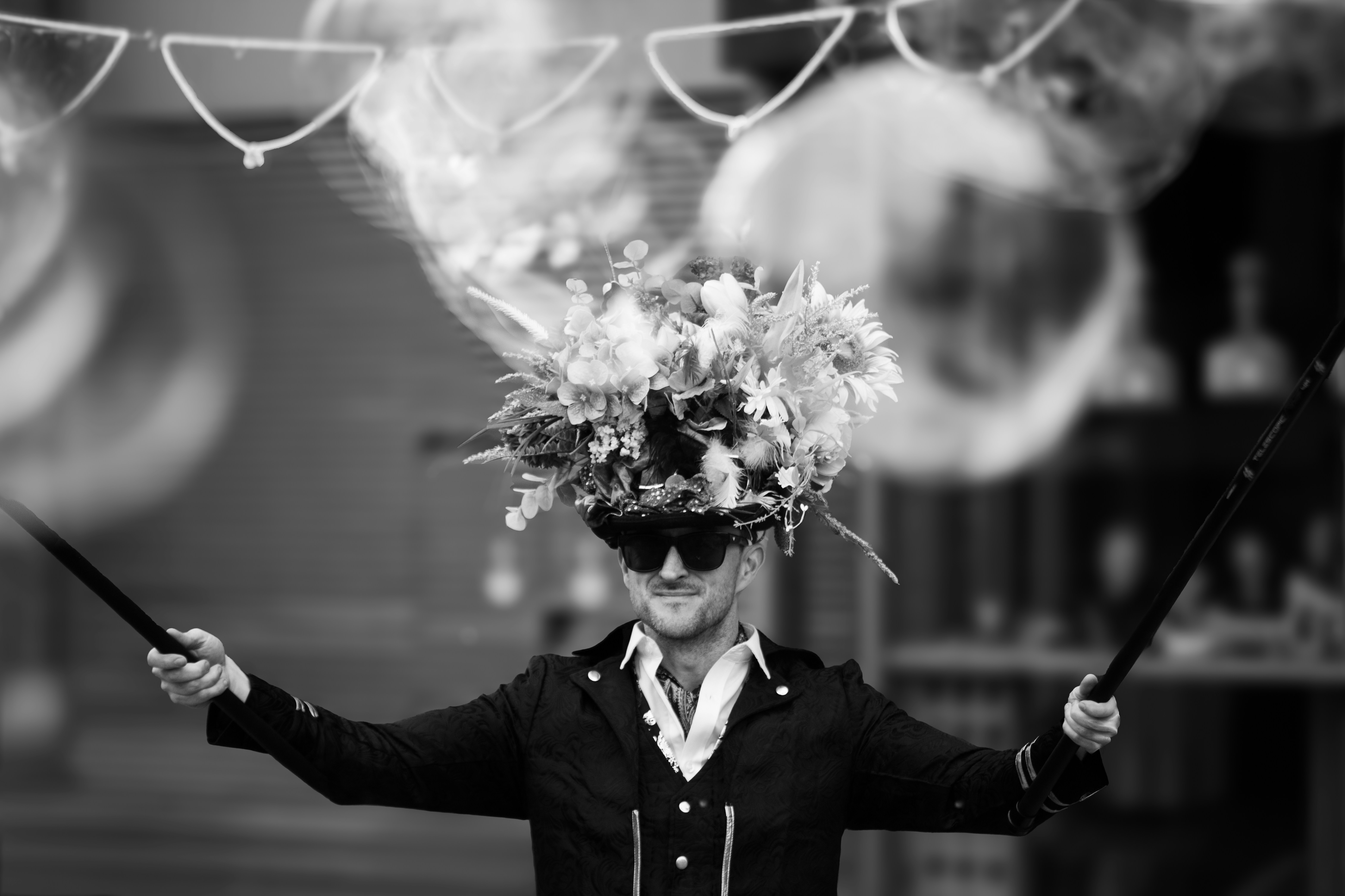 A monochrome portrait of a performer with a dramatic floral crown, arms extended holding two wands, against a blurred backdrop. This image emphasizes the subject's headpiece and posture, presenting a stylized, artistic moment.