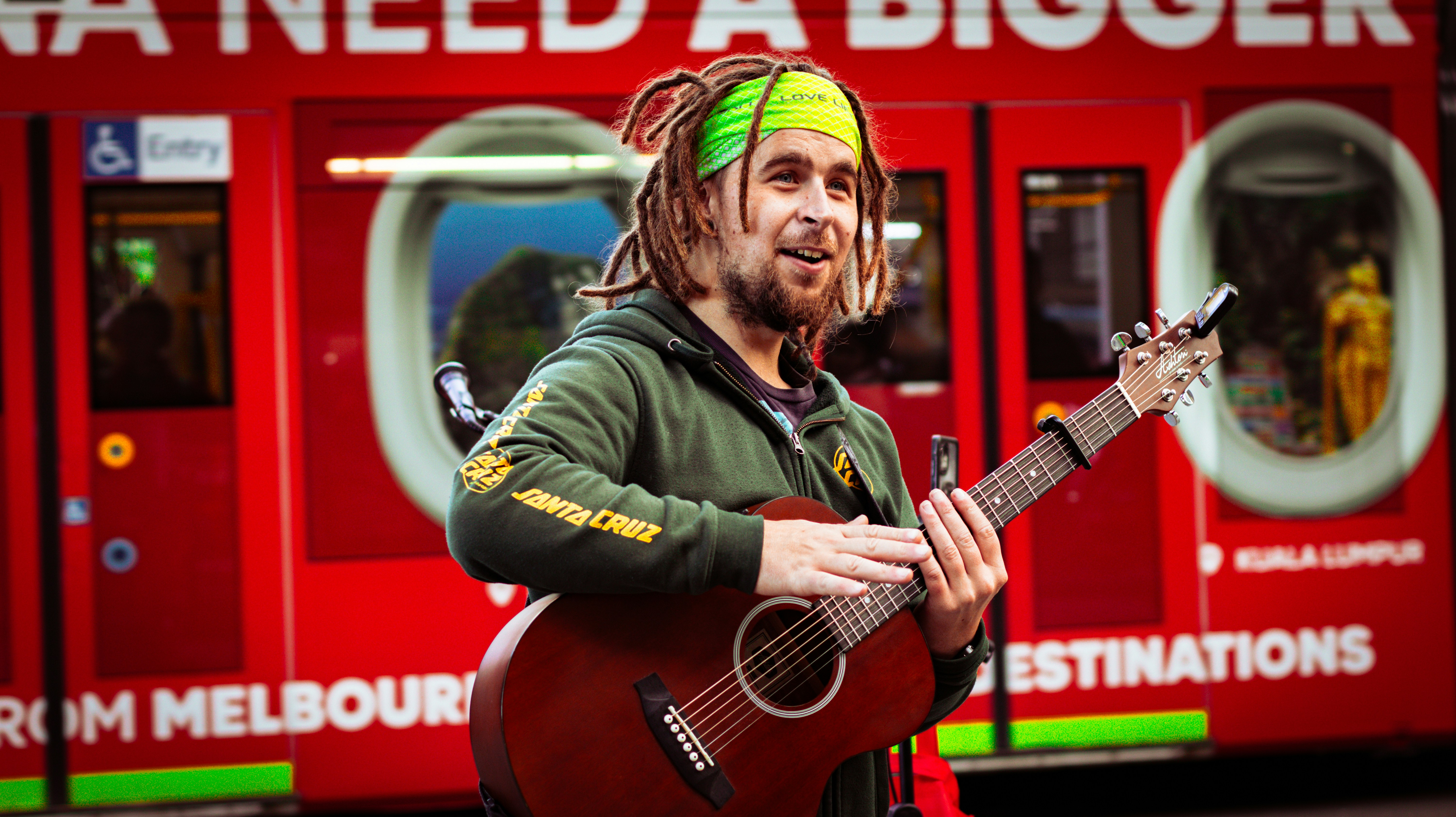 Busker with Guitar in Front of Red Transit Bus