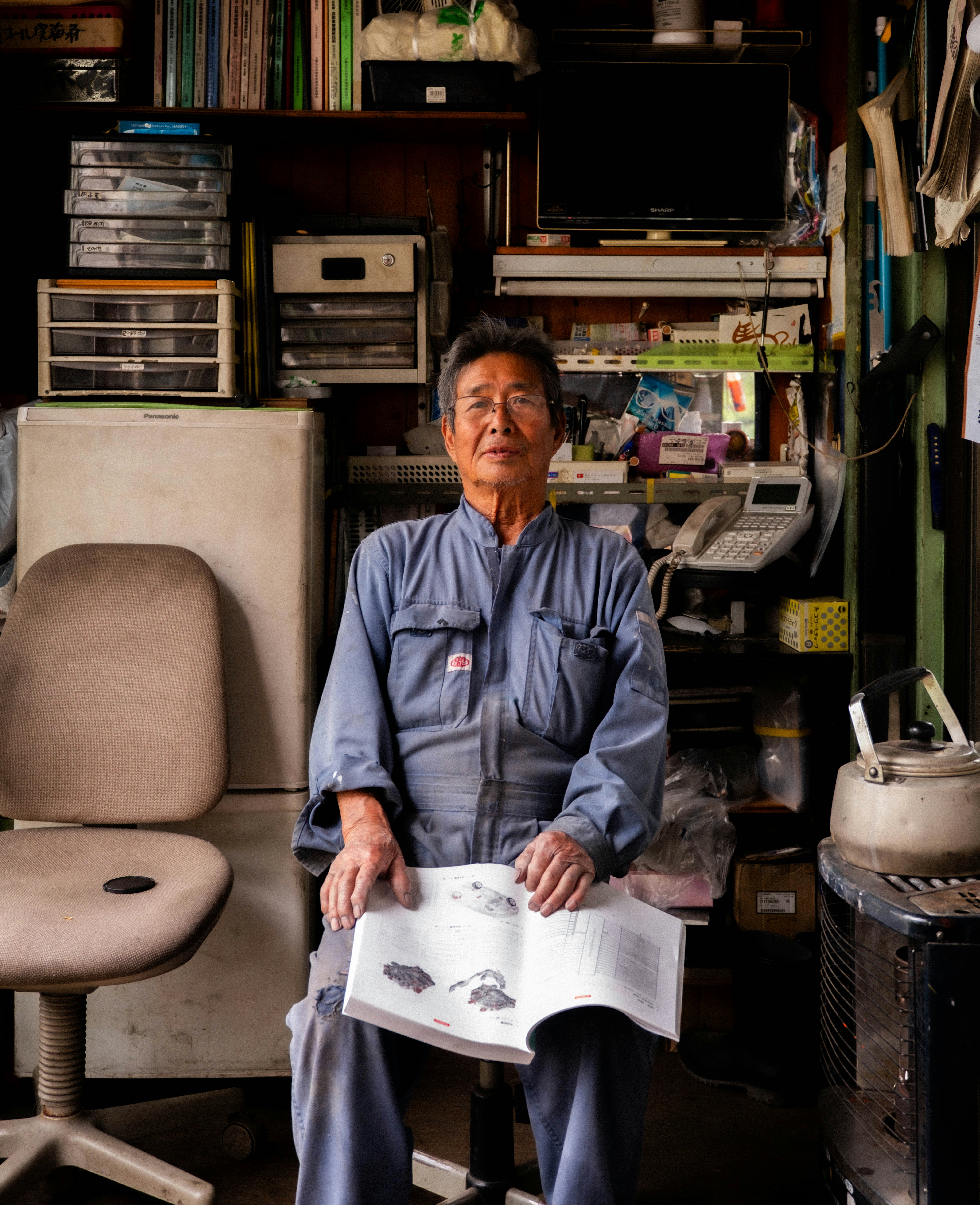 An elderly man in blue workwear sits center in a cluttered workshop, reading a magazine as various tools and equipment surround him. This is a candid, real-world interior scene showing everyday labor and concentration.