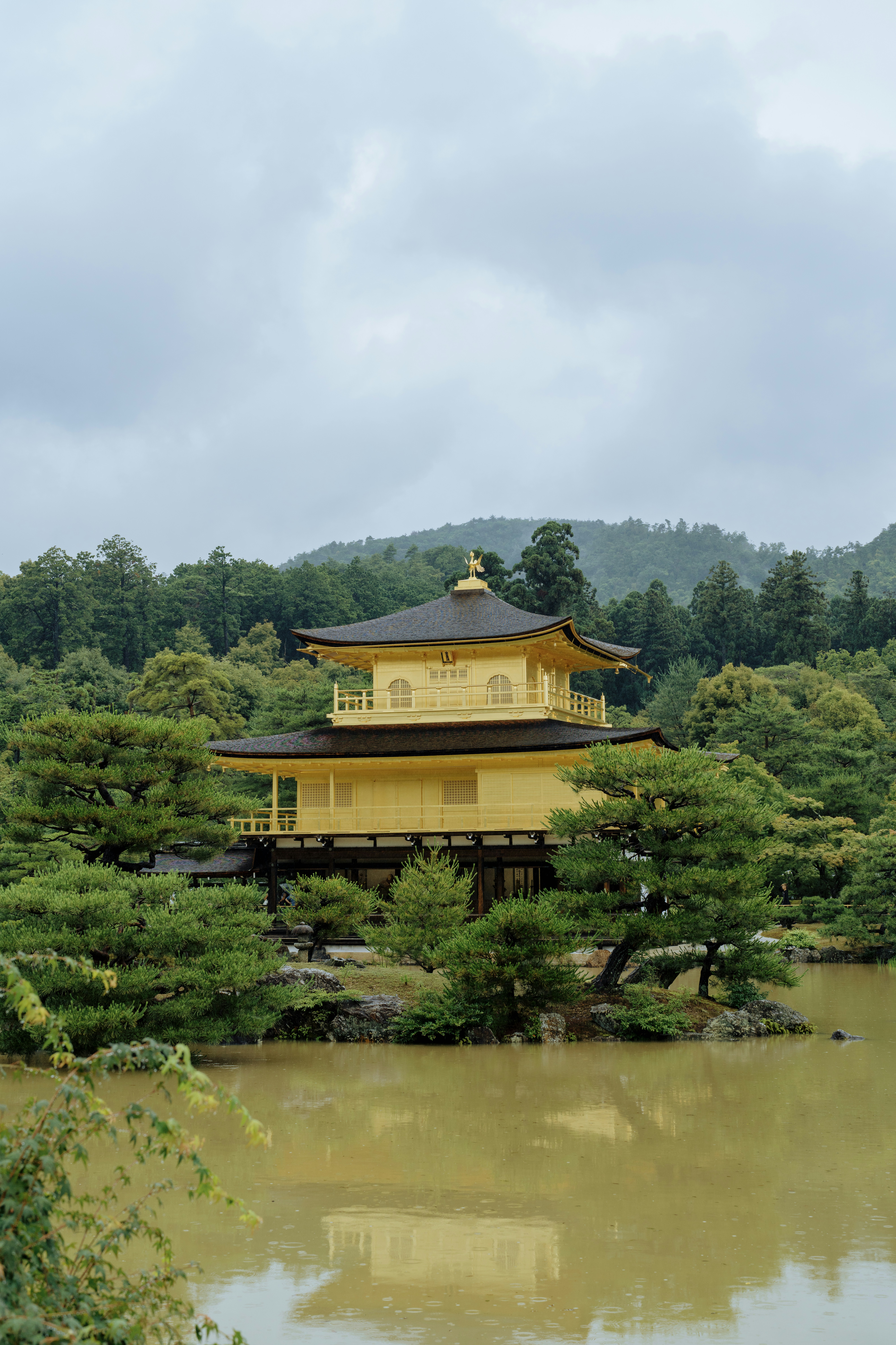 Golden pavilion rising from a tranquil pond with surrounding trees and soft sky hues, captured as a tranquil architectural scene.