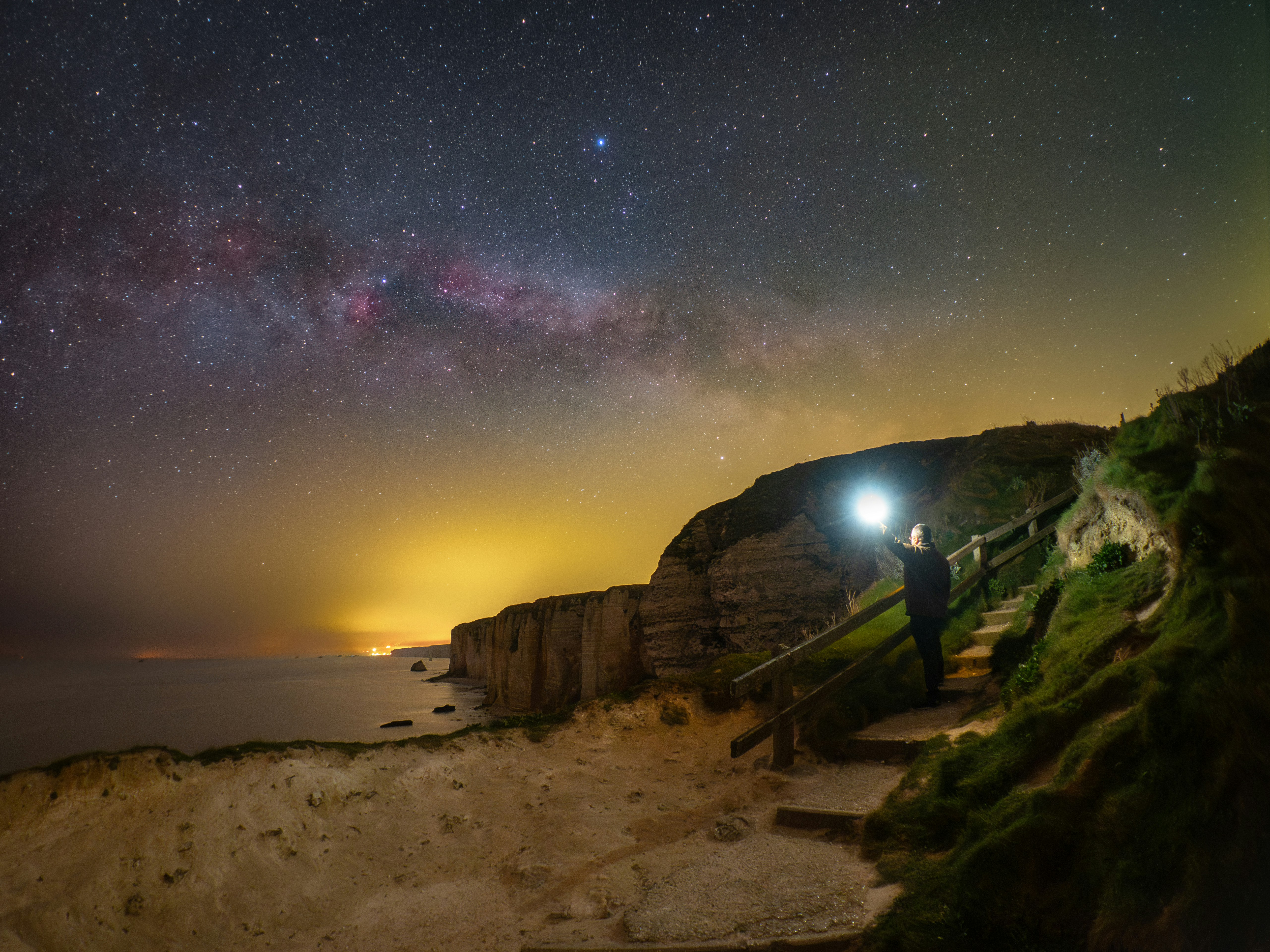 Night under the Milky Way: a lone explorer Illuminates the cliffside trail