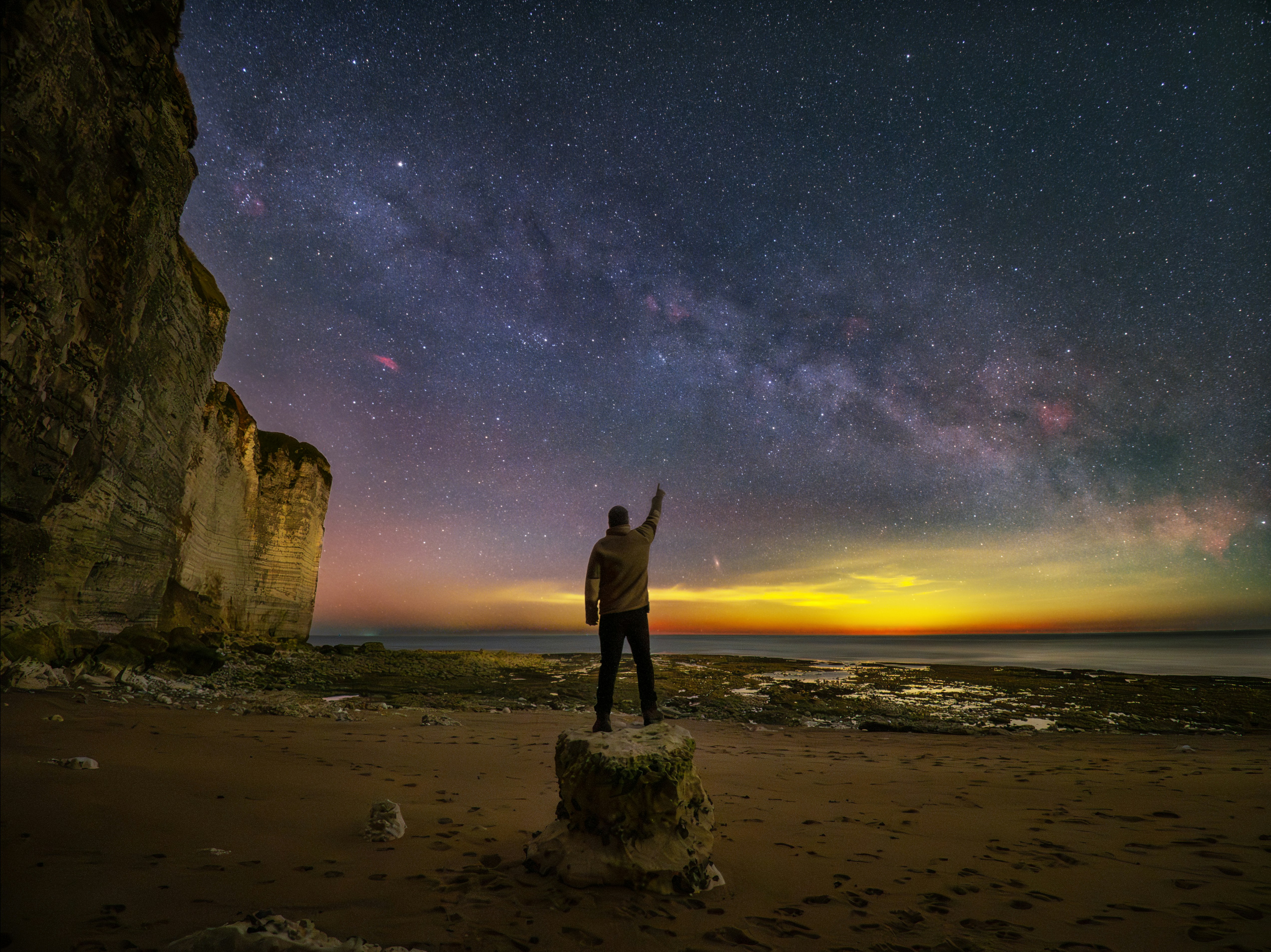 Man on beach points to milky way and glowing horizon.