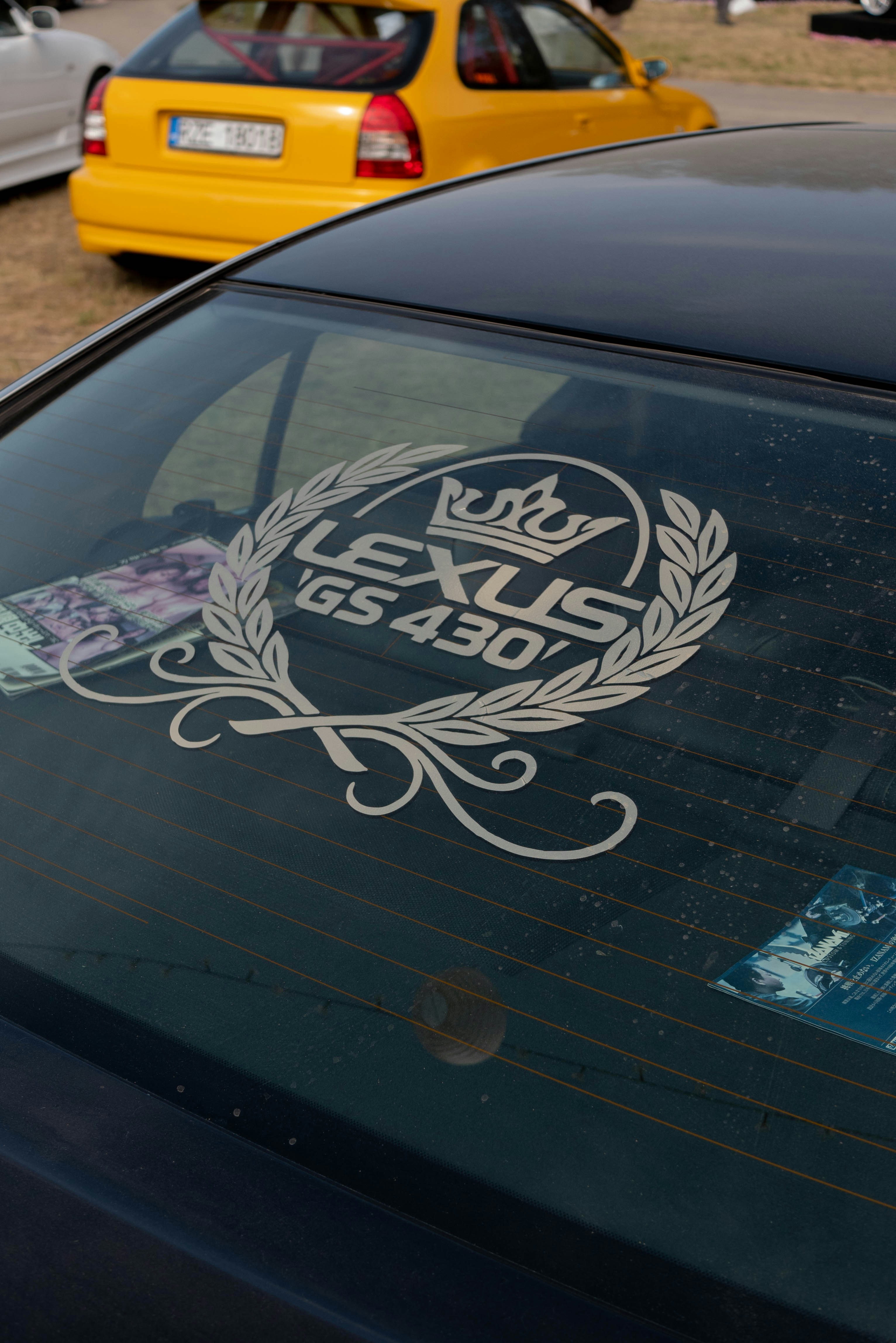Rear windshield decal reads 'LEXUS GS 430' framed by a laurel emblem on a dark car; yellow hatchback and another vehicle visible in the background.