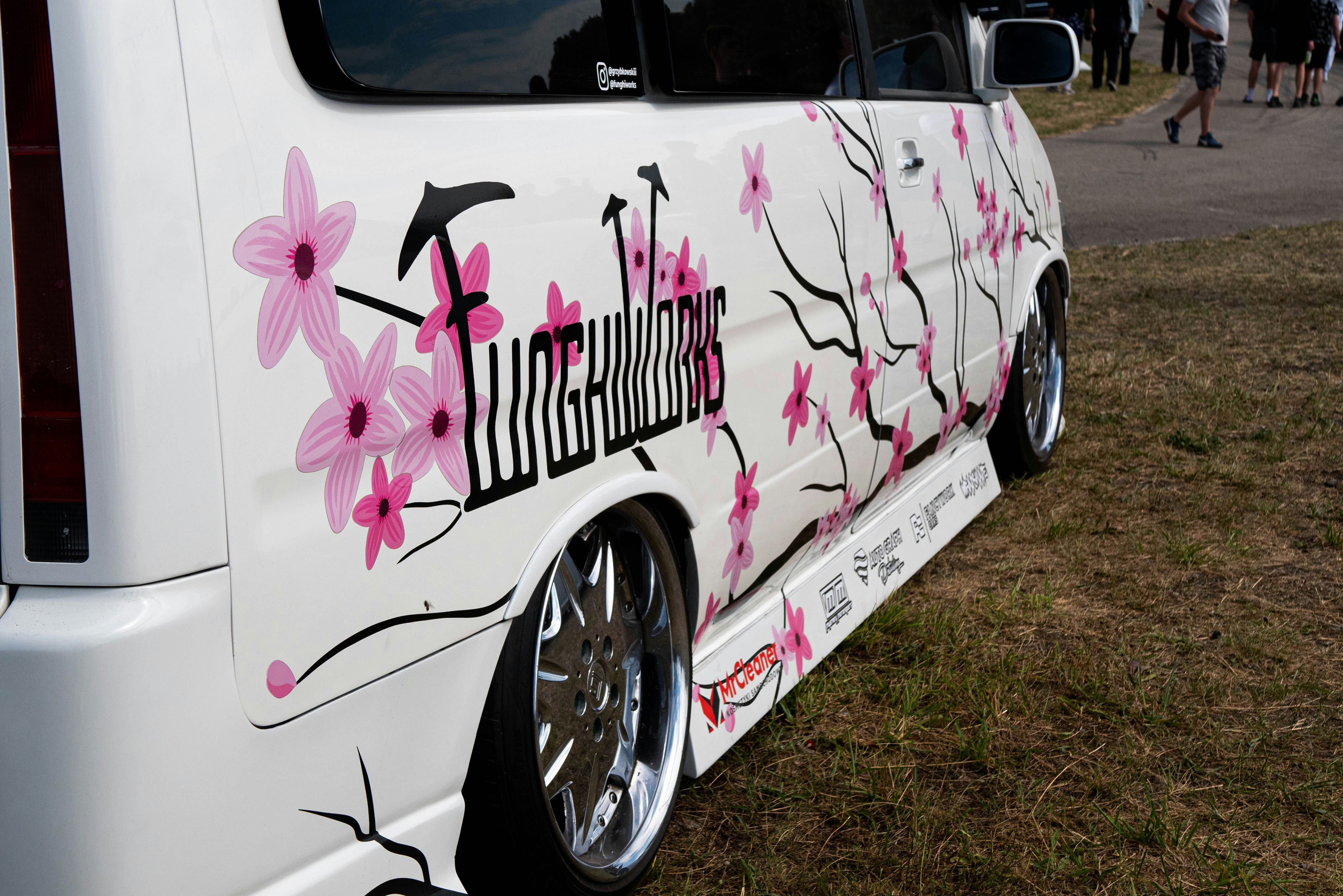 White van with pink cherry blossom graphics and bold black lettering parked on grass; highlights decorative wrap and chrome wheels.