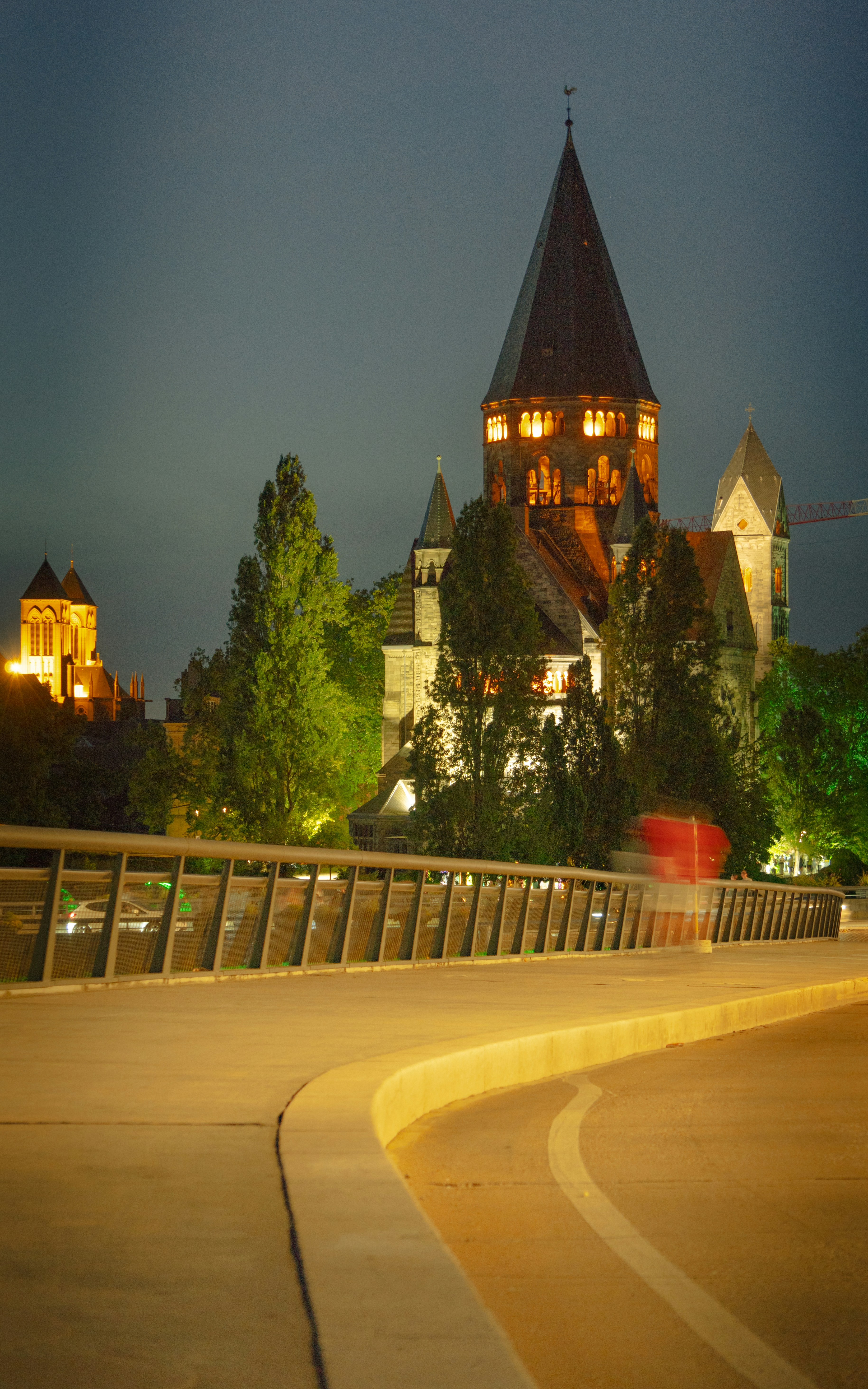 Night scene of a historic church/castle complex lit from within, seen over a curved riverside walkway; soft motion blur on a passing figure adds a sense of liveliness. Likely a cityscape with a logo-like lighting on the structure.