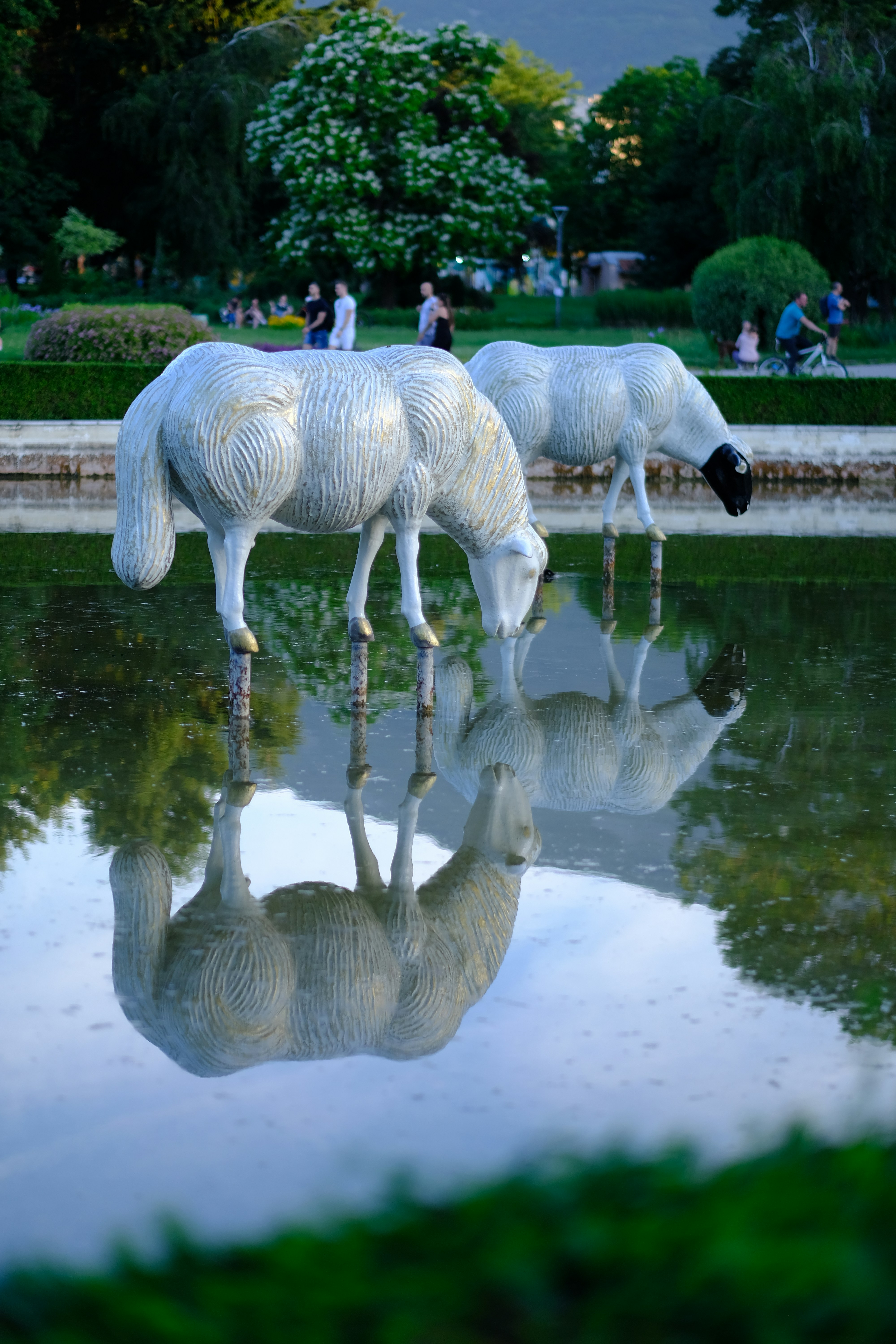 White horse sculptures graze in a shallow reflective pool with a park and visitors in the background.