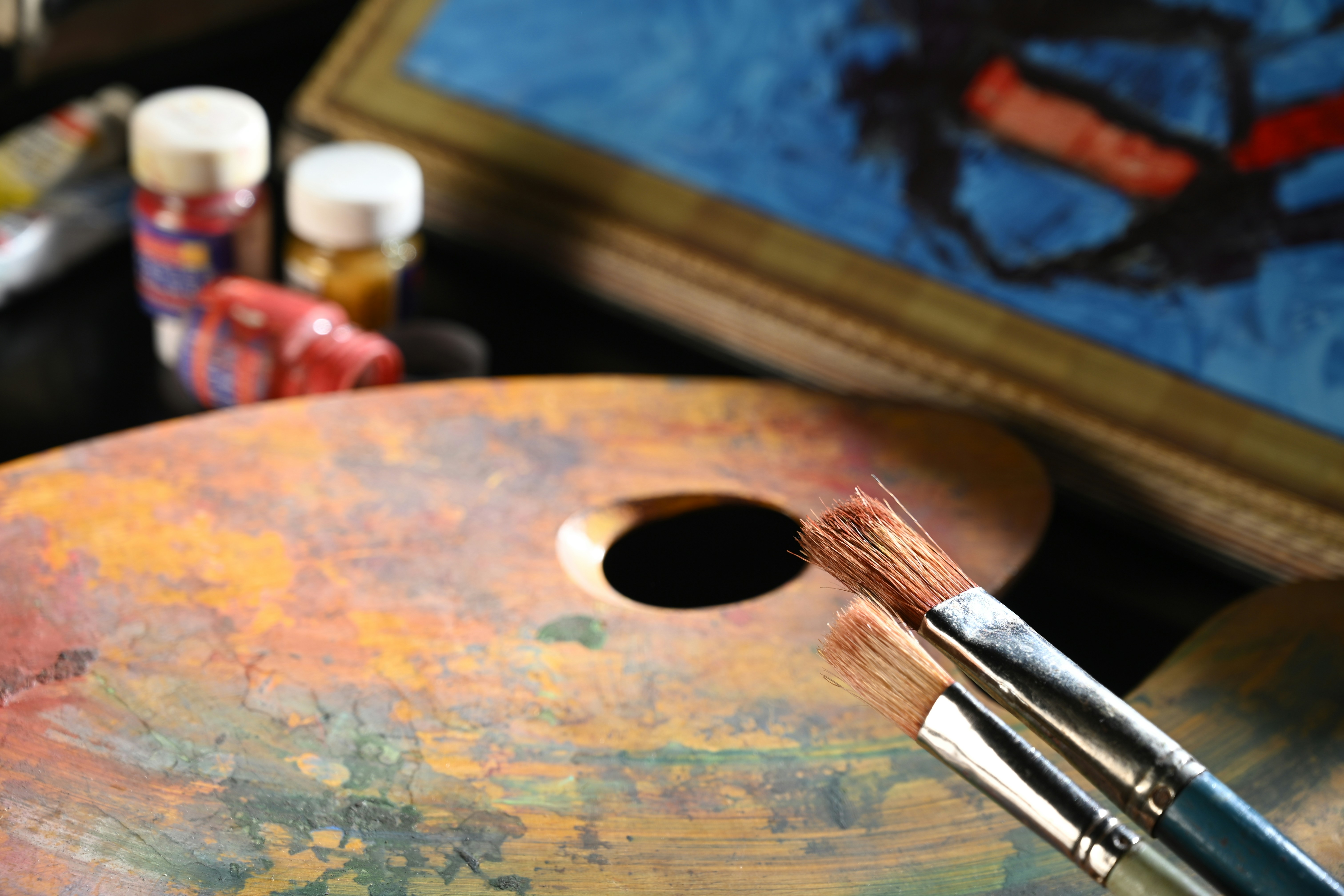 Close-up of a painter's palette with brushes resting nearby, acrylic paints in the background and a canvas in the back; a still life of art supplies focusing on texture and color. This is a photographed setup with studio lighting.