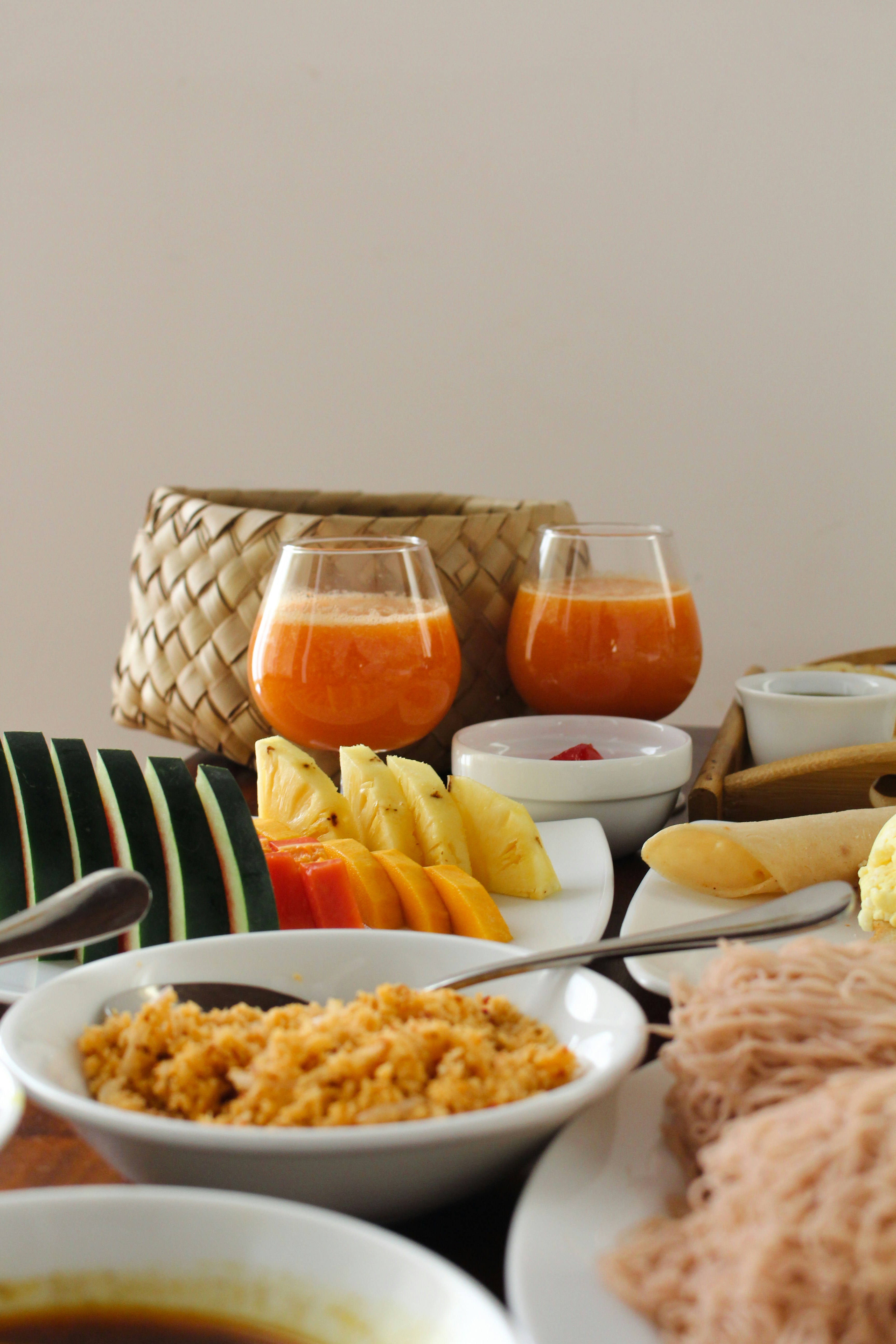 Vibrant fruit platter, noodles, and juices laid out on a dining table for a casual meal setup. A woven basket and assorted bowls frame the spread.