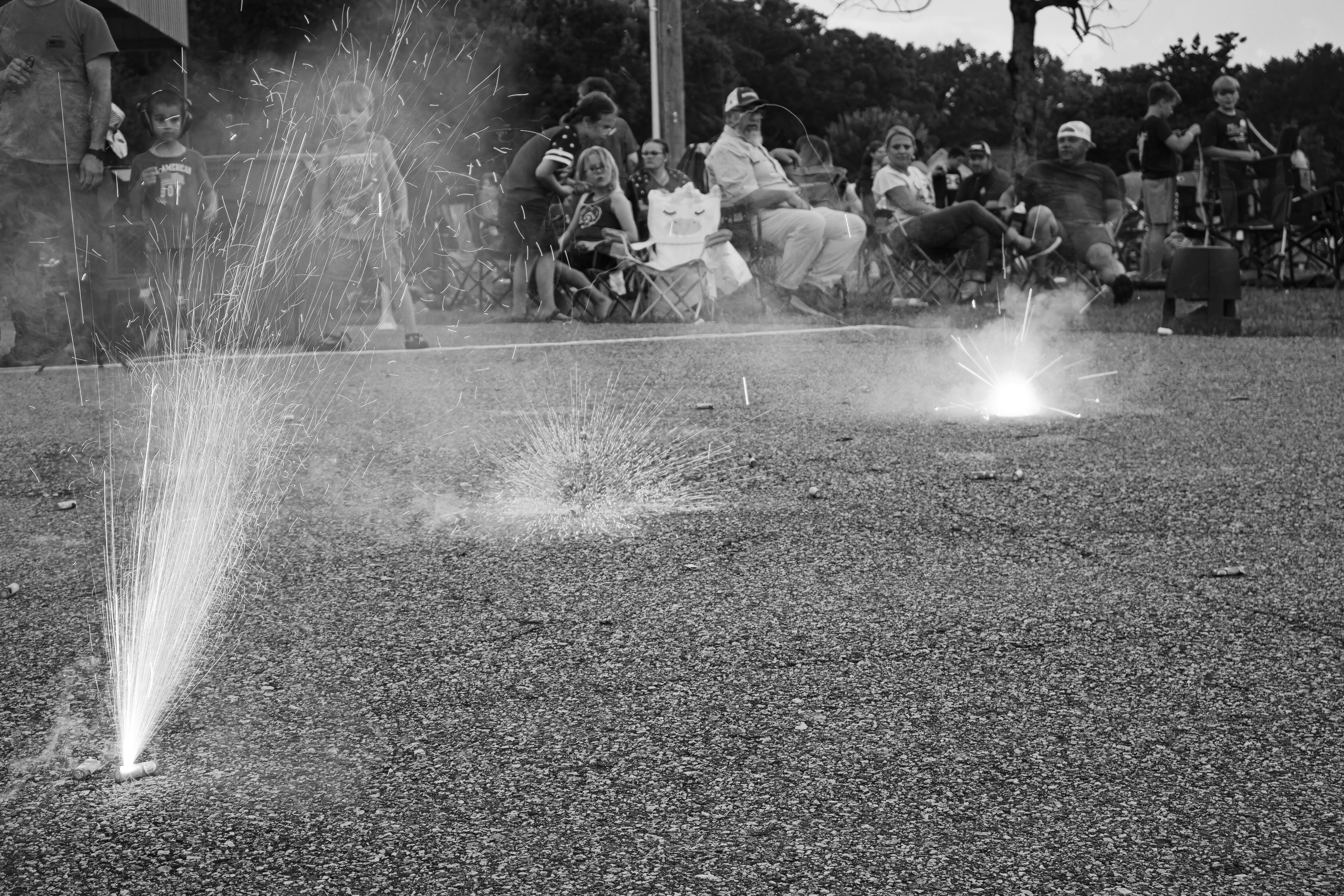 Close-range view of ground-based fireworks erupting with bright sparks, spectators seated along a park lawn in the background; a candid moment captured to convey celebration and communal viewing.