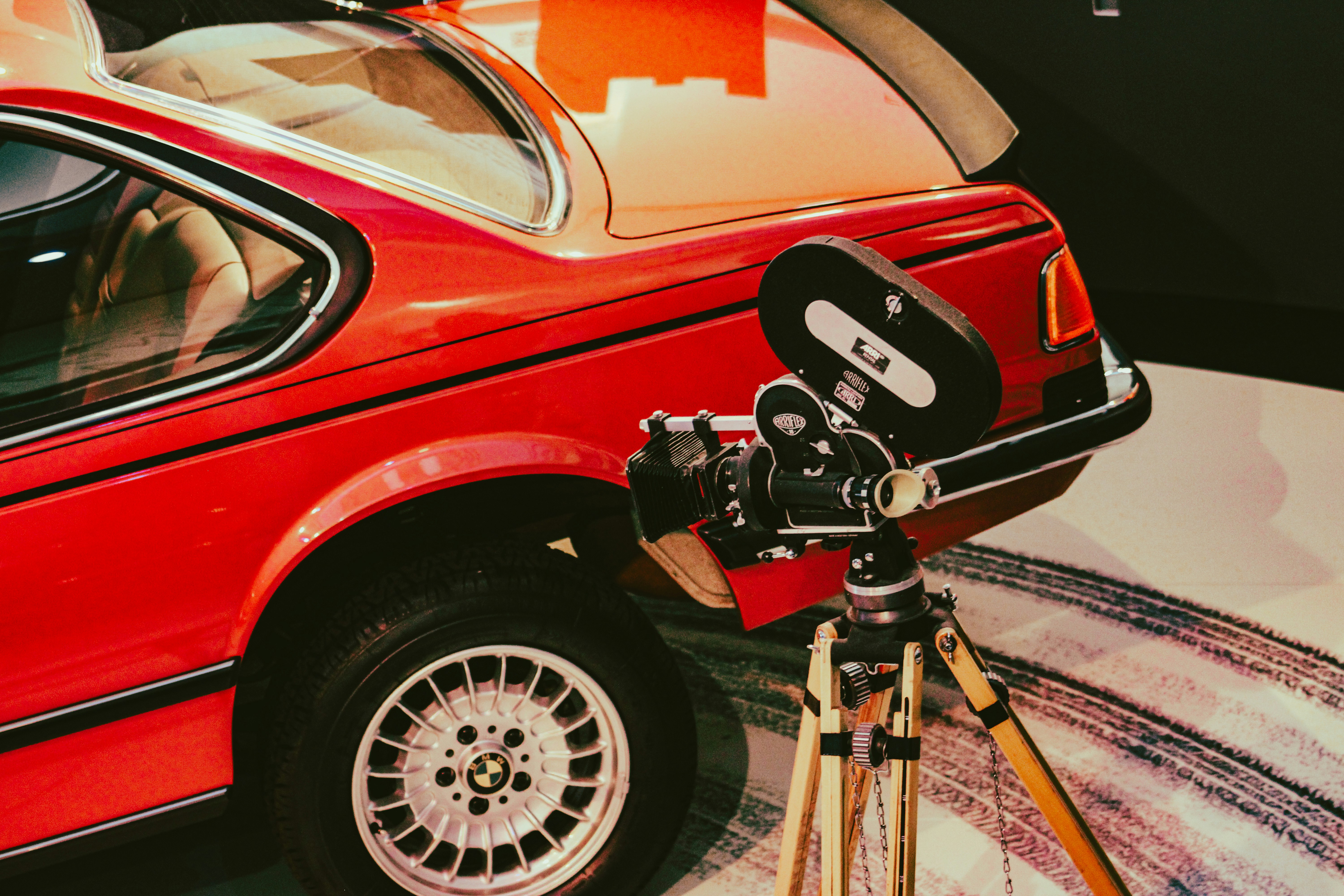 Red vintage car with a classic rangefinder-style camera on a tripod set against a dark backdrop; a detailed view emphasizes the car’s rear and wheel as the focal composition. This is a staged photographic scene.