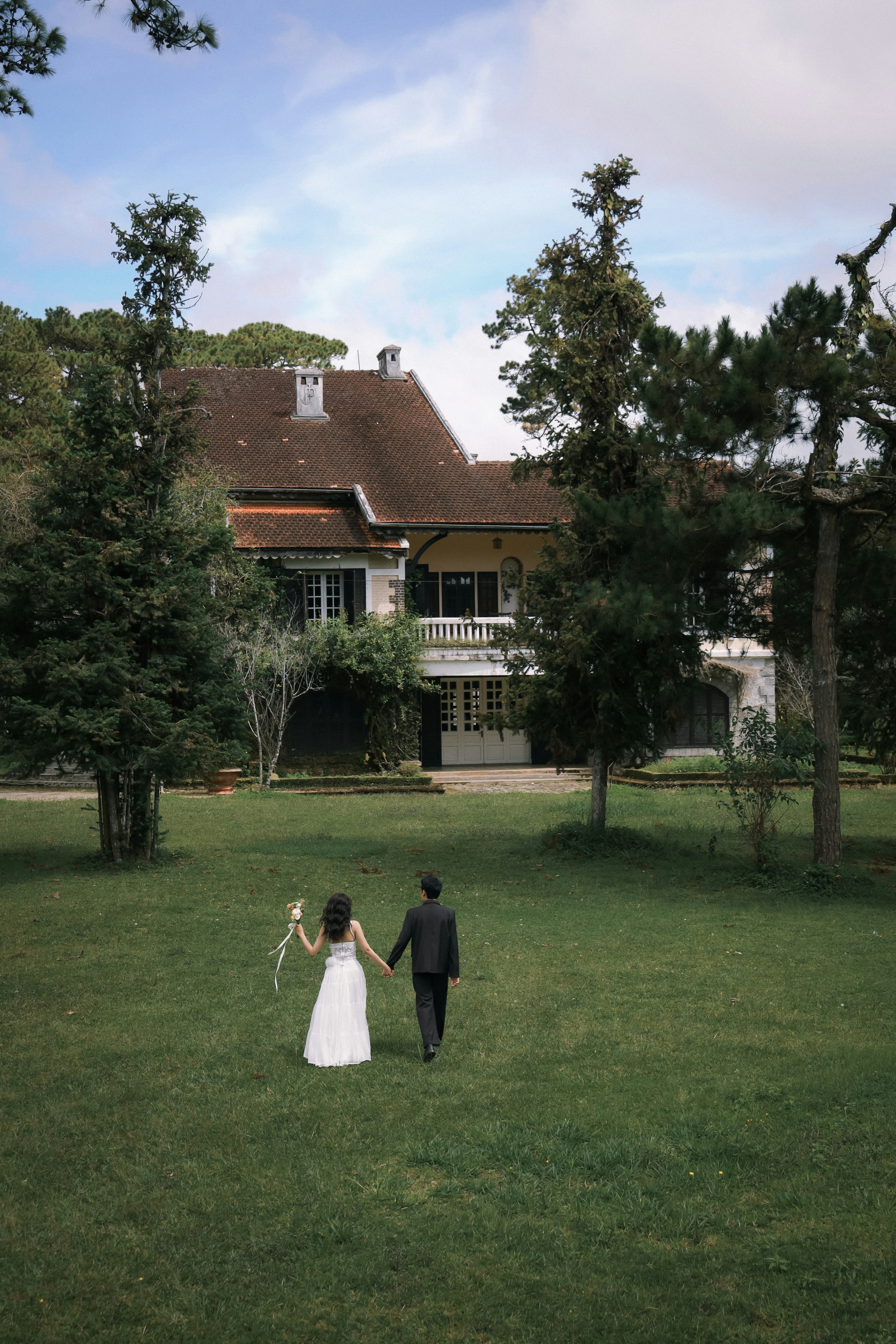 Bride and groom walk hand-in-hand across a manicured lawn toward a stately house, framed by tall pines in a serene outdoor setting.