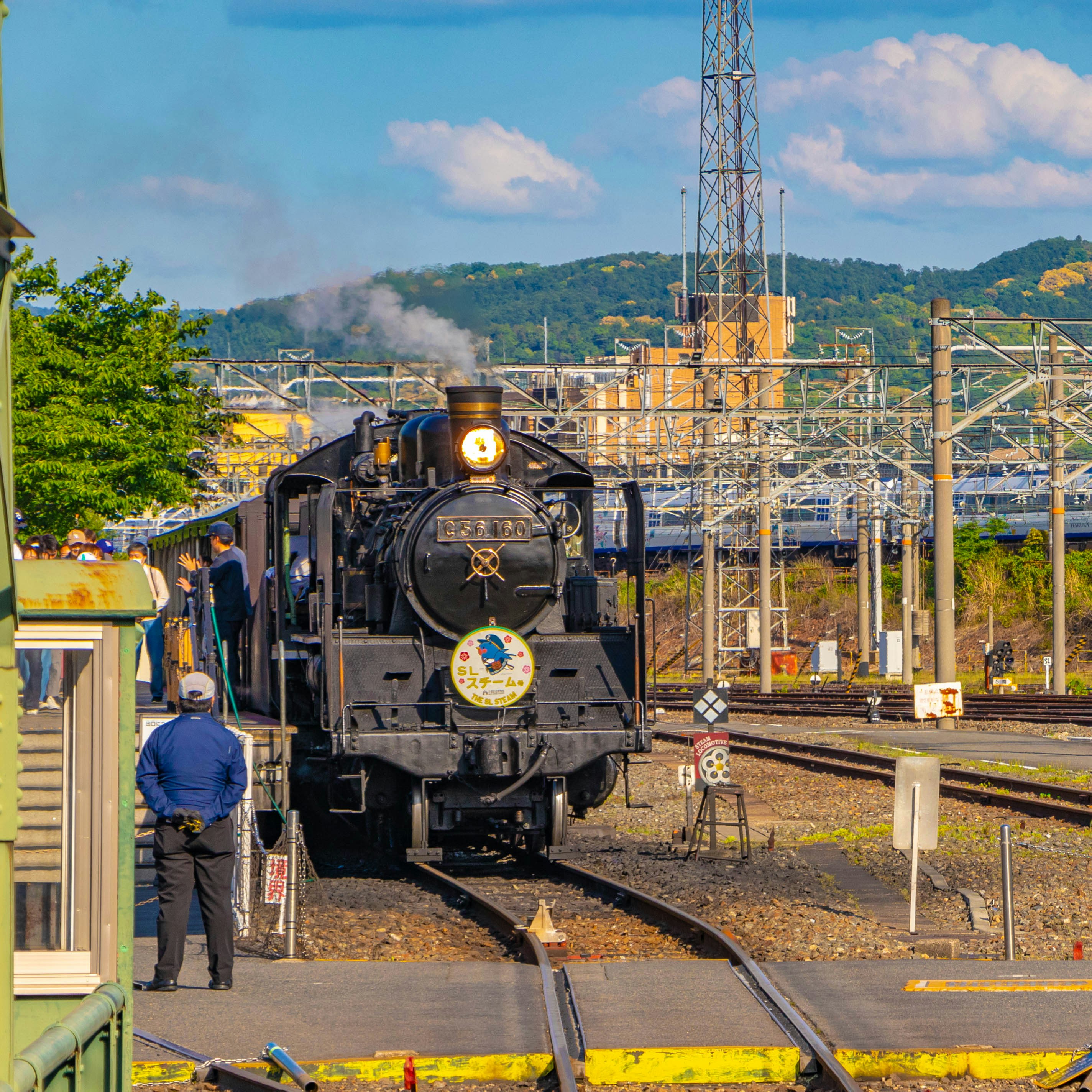 Steam locomotive facing the yard with crewman nearby; foreground rails and signal equipment anchor the scene in a busy rail depot.
