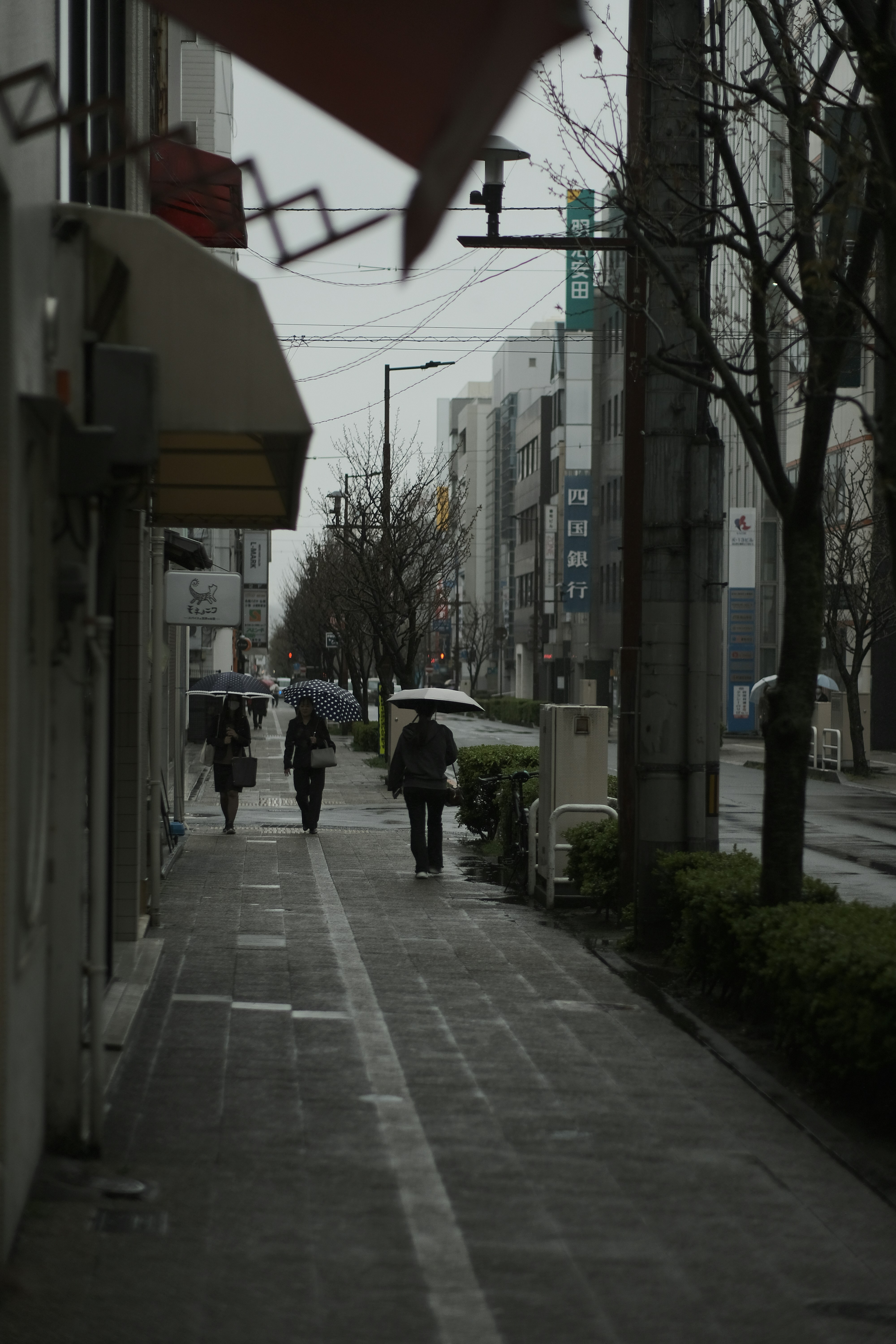 Rainy sidewalk in a city: pedestrians with umbrellas under overhangs