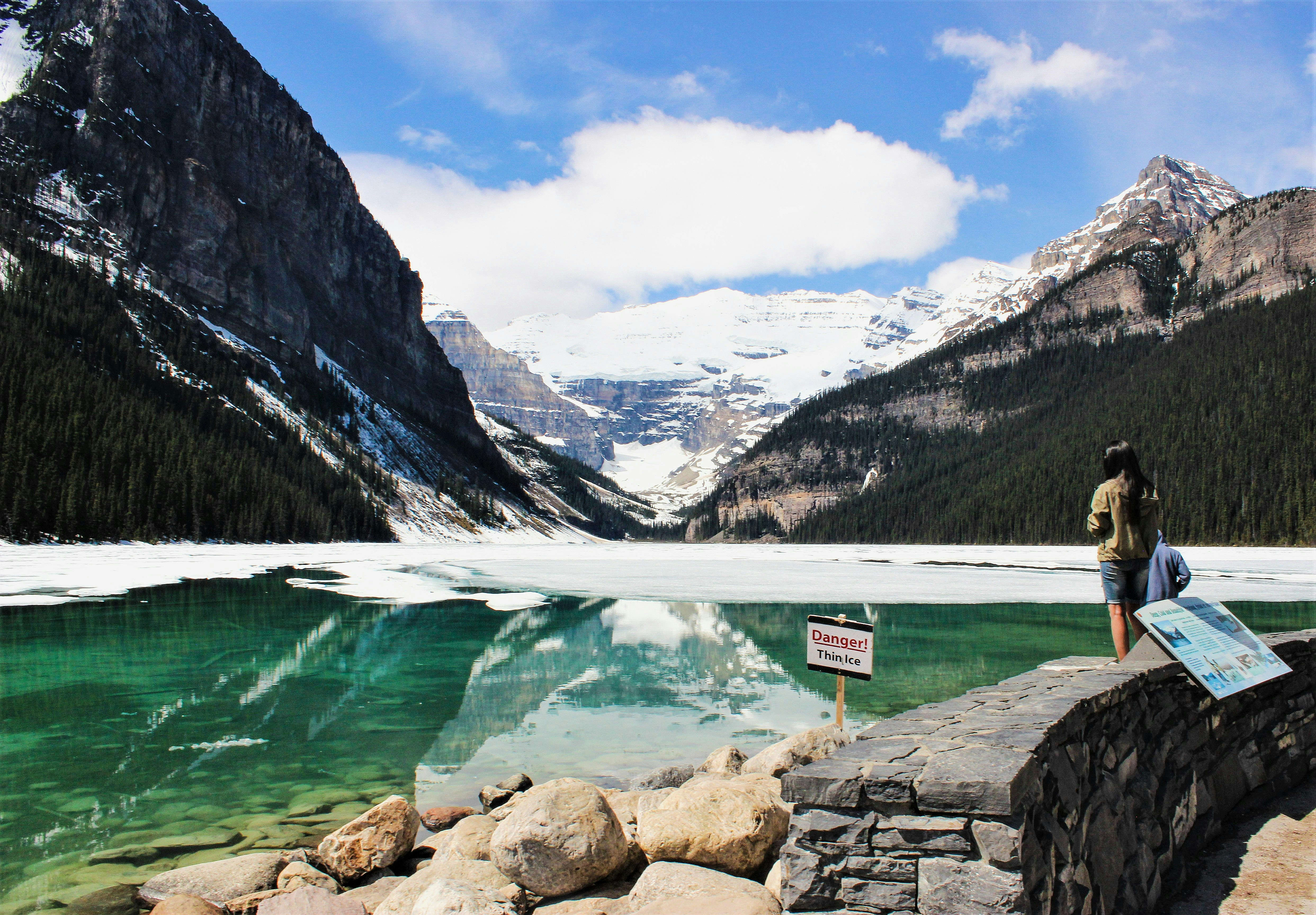 A stunning spring view of Lake Louise in Banff National Park, Alberta, Canada, where turquoise glacial water emerges through melting ice beneath towering snow-covered Rocky Mountains. Visitors admire the breathtaking alpine scenery as reflections shimmer across the crystal-clear lake, capturing the beauty of Canada's iconic wilderness.