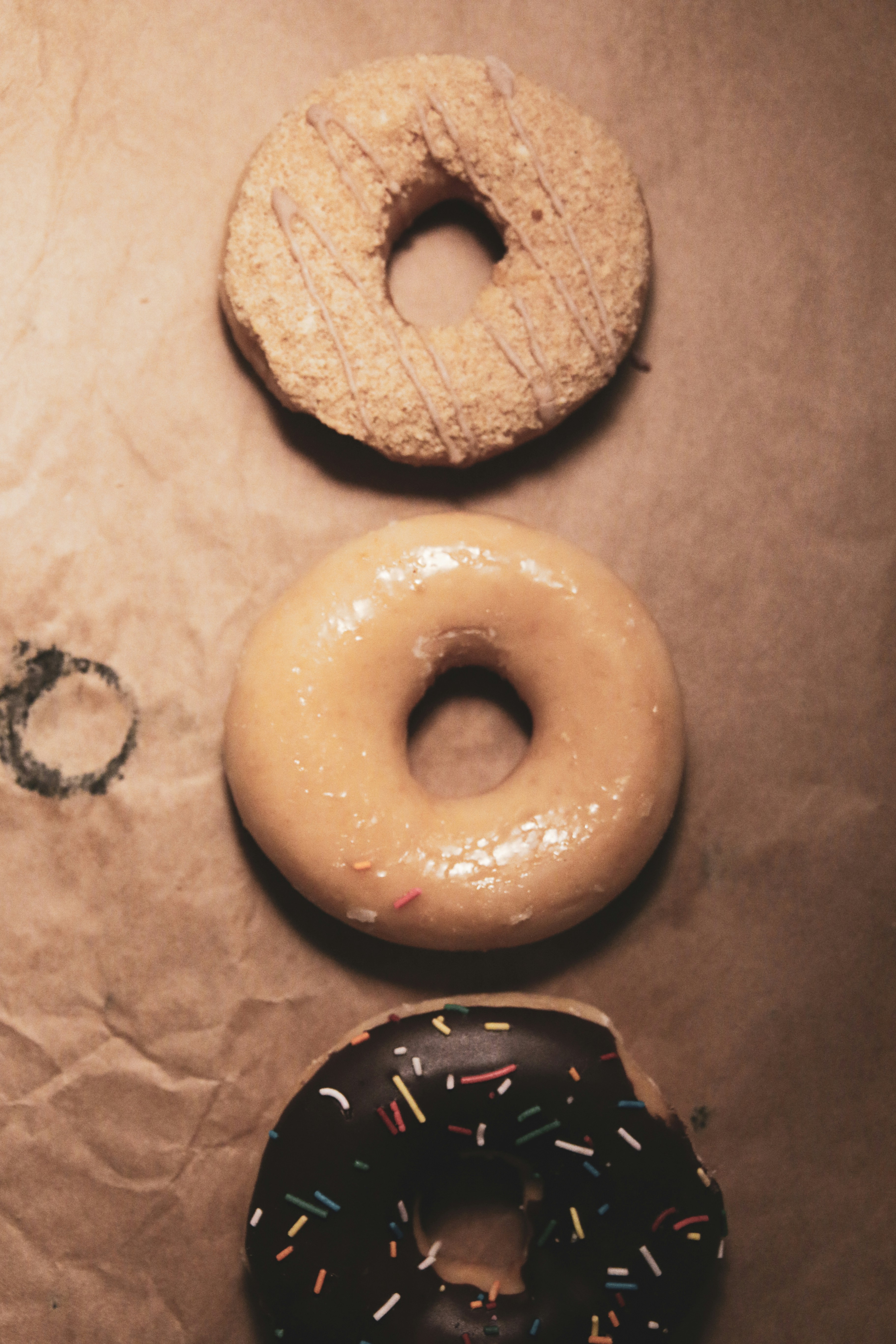 Three donuts arranged vertically on parchment, from toasted crumb to glossy chocolate with sprinkles, inviting a bite. A kitchen still-life focusing on glaze textures and sugars.