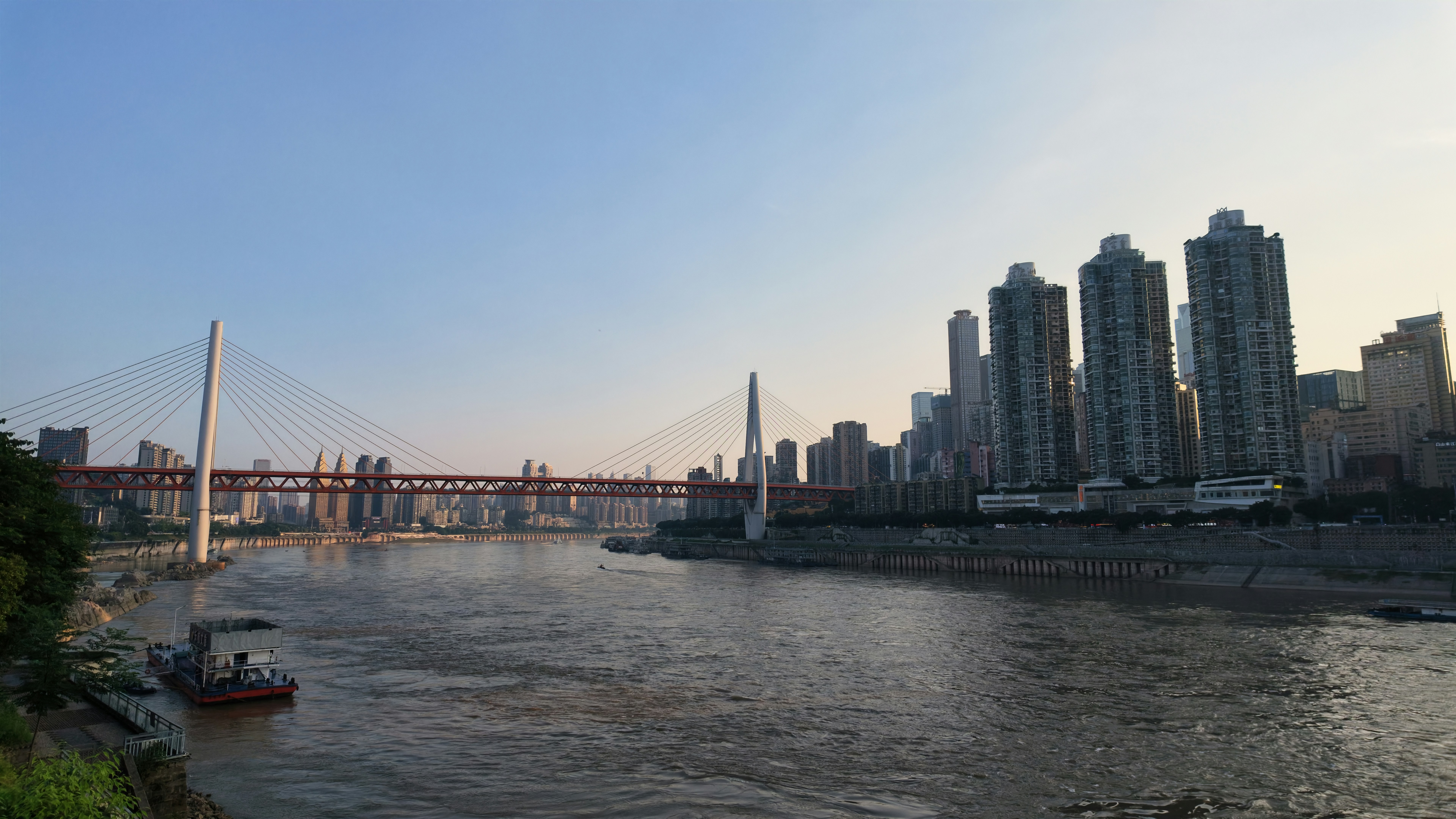 City riverfront with a cable-stayed bridge and cluster of high-rise buildings along the bank; calm water reflects the skyline, captured from the riverbank.