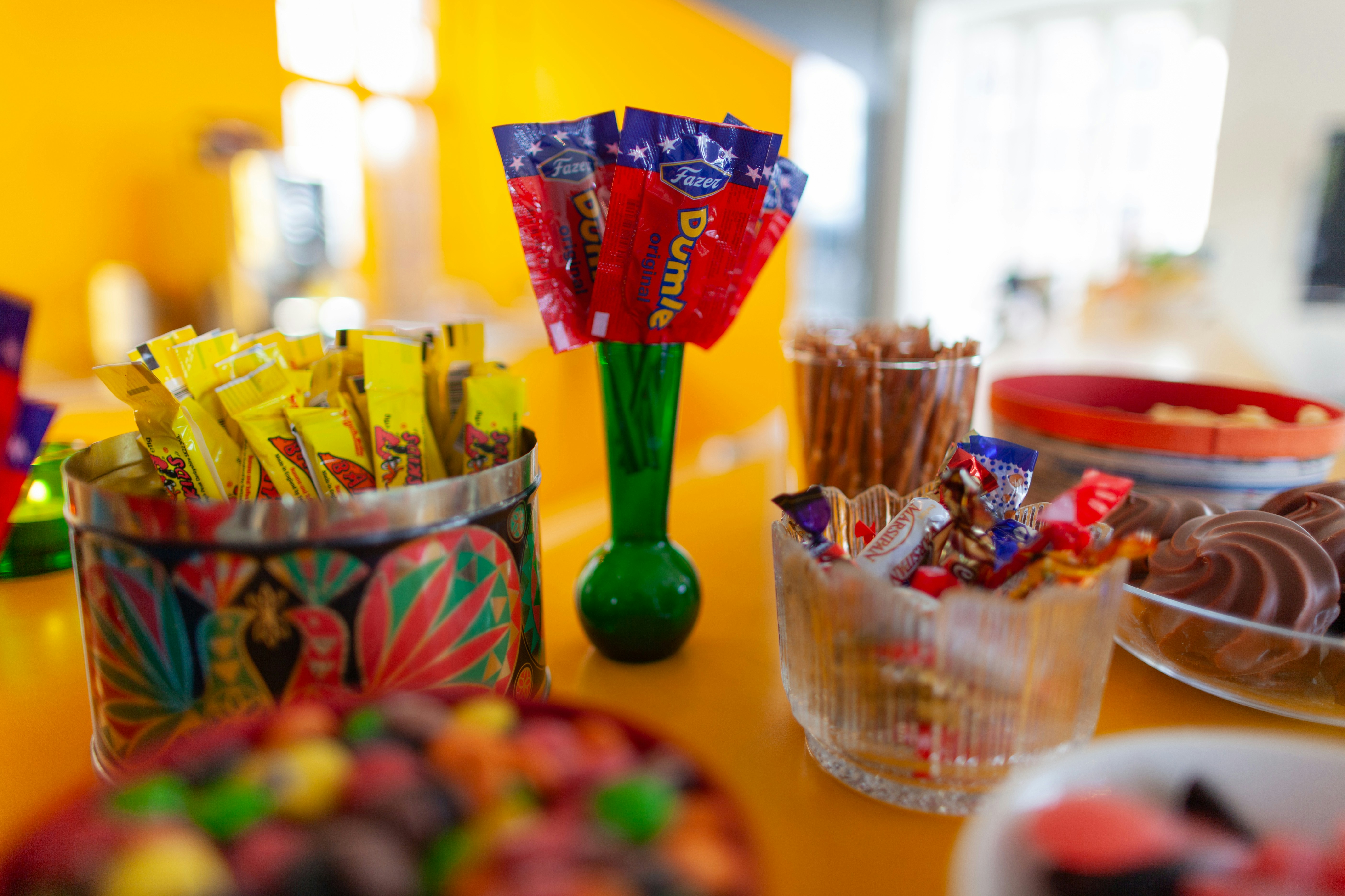 Assorted candies in colorful containers on a sunny, yellow kitchen counter; a festive spread with wrappers and a variety of sweets presenting a vibrant still-life scene.