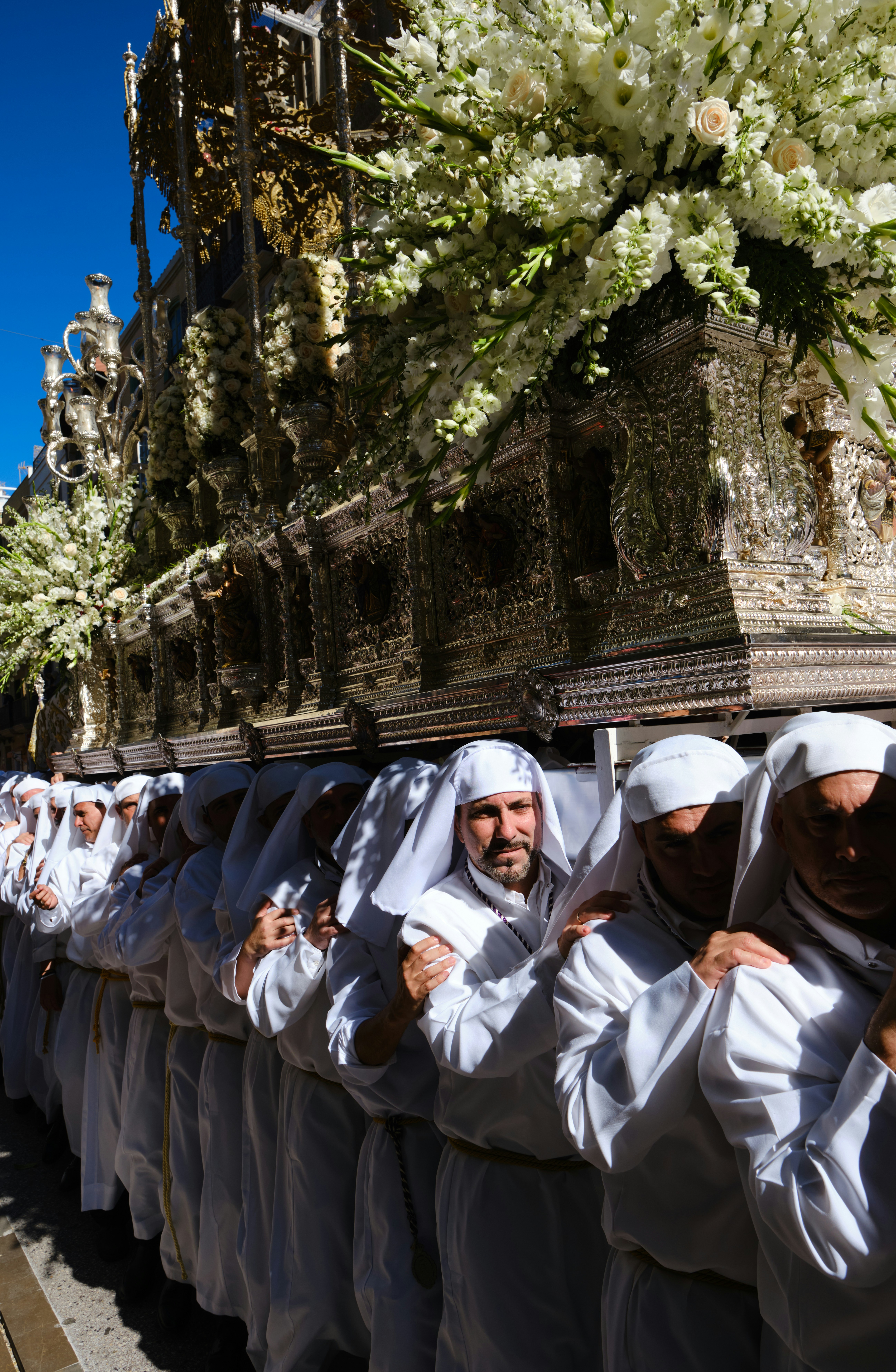 White-robed mourners carry a floral reliquary through a sunlit urban avenue