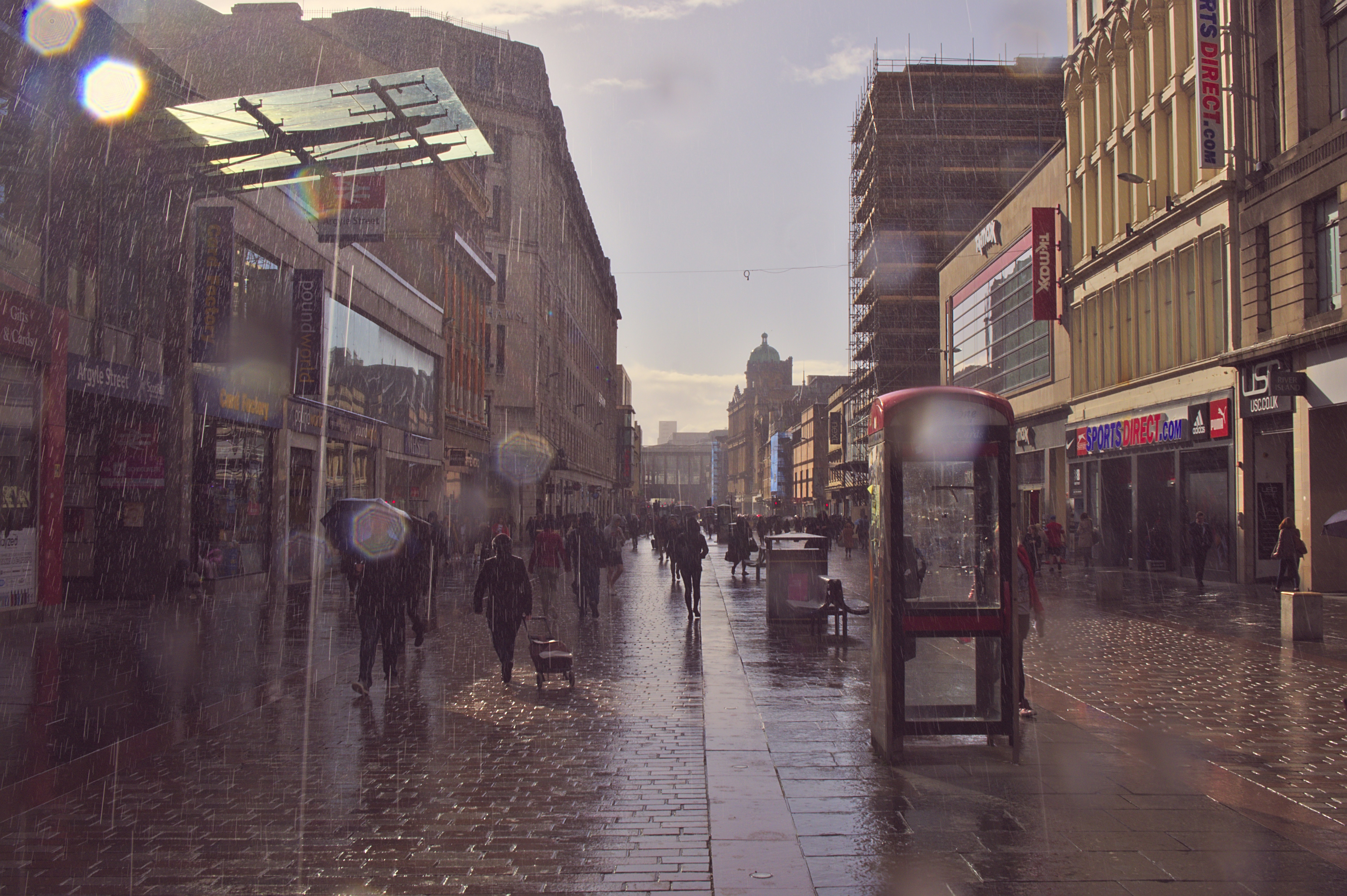 Rain-soaked city street with pedestrians, a red bus stop, and reflections on the wet cobblestones under overcast skies.