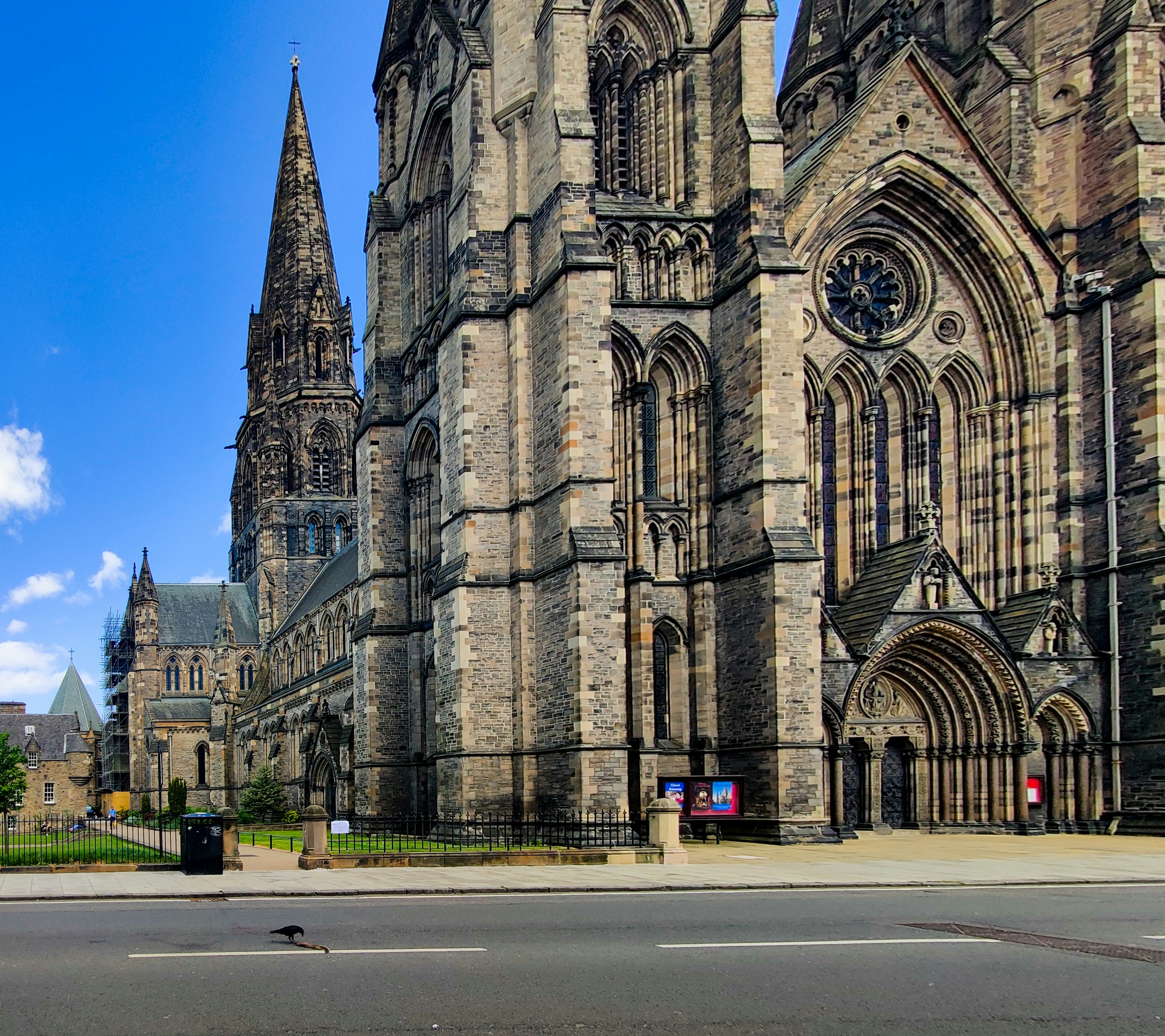 Gothic cathedral facade dominates a quiet urban street, with a stone exterior, arched doorways, and a small roadside scene including a digital display and a lone bird on the pavement.