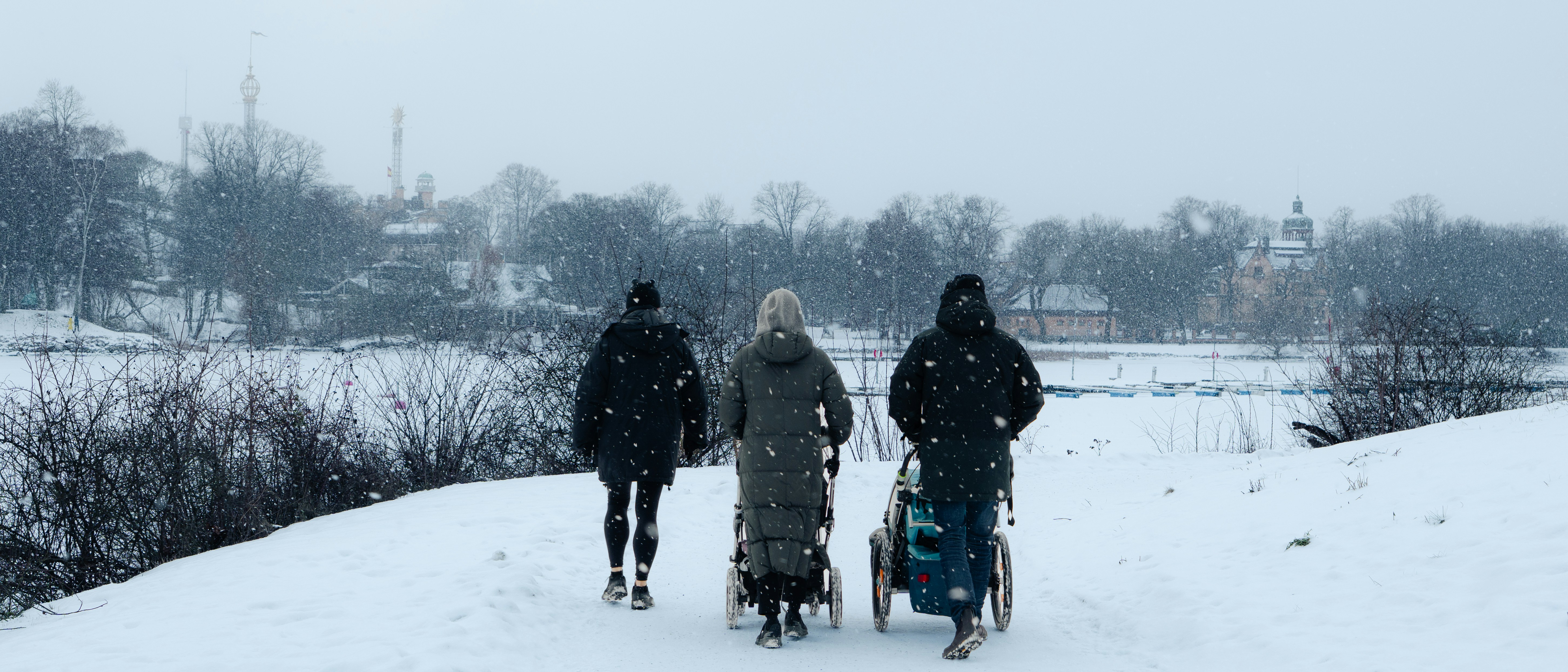 Three figures push strollers along a snowy riverside path, silhouettes against a pale, snow-laden landscape with distant buildings under overcast skies. A calm, wintry scene with a focus on contrast between dark coats and white snow.