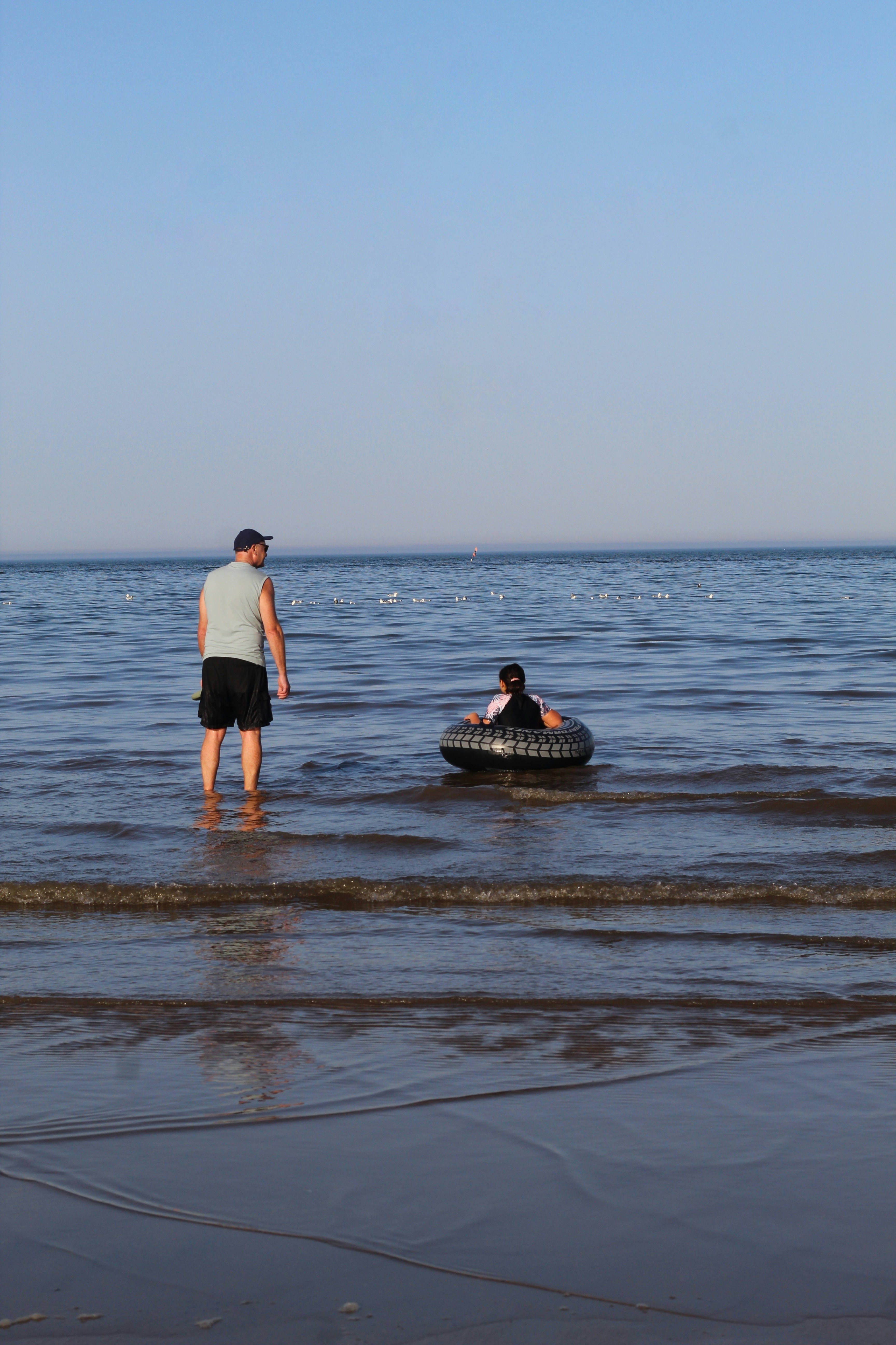 Two figures, a man standing in shallow water and a person in an inner tube, gaze toward the calm horizon on a clear day at the beach. The scene captures a relaxed moment of seaside play. (Photo, not a painting or illustration.)
