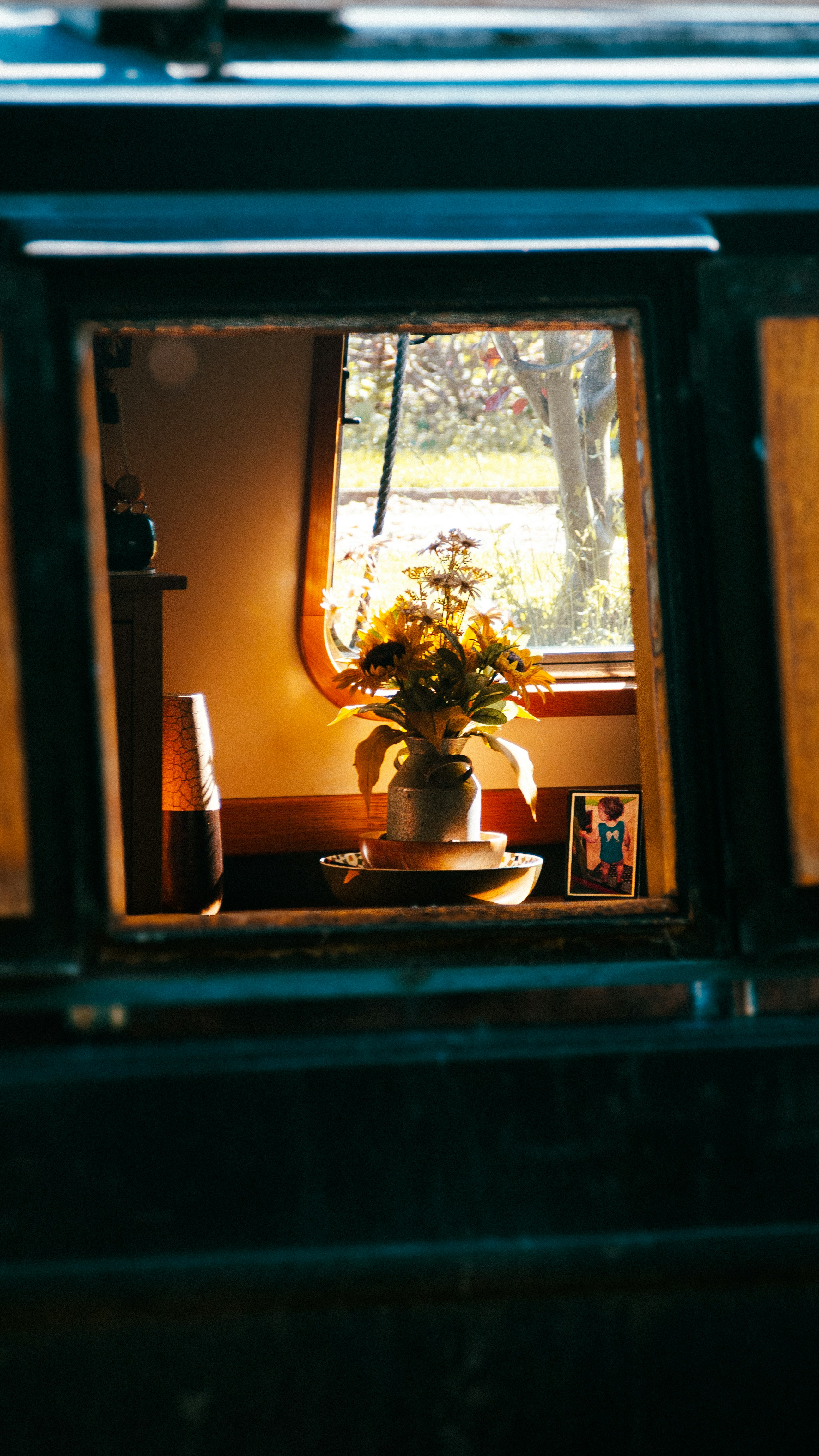 A sunlit interior scene framed by a window, featuring a vase of flowers on a table with a small framed photo beside it, viewed through a dark surrounding frame.