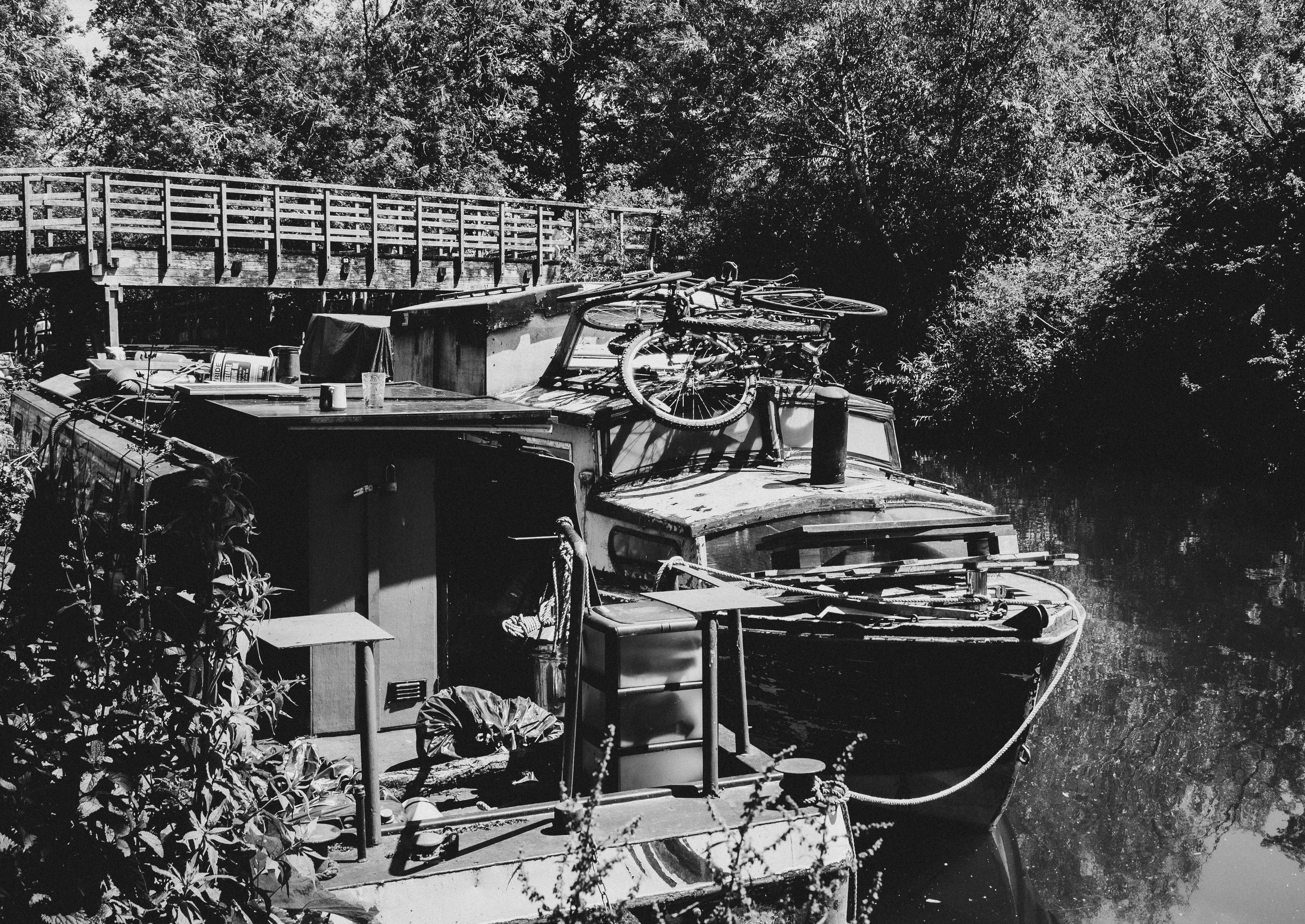 Monochrome scene of a weathered houseboat moored beside a wooden walkway, with bicycles strapped to the roof and overgrown vegetation lining the hull.