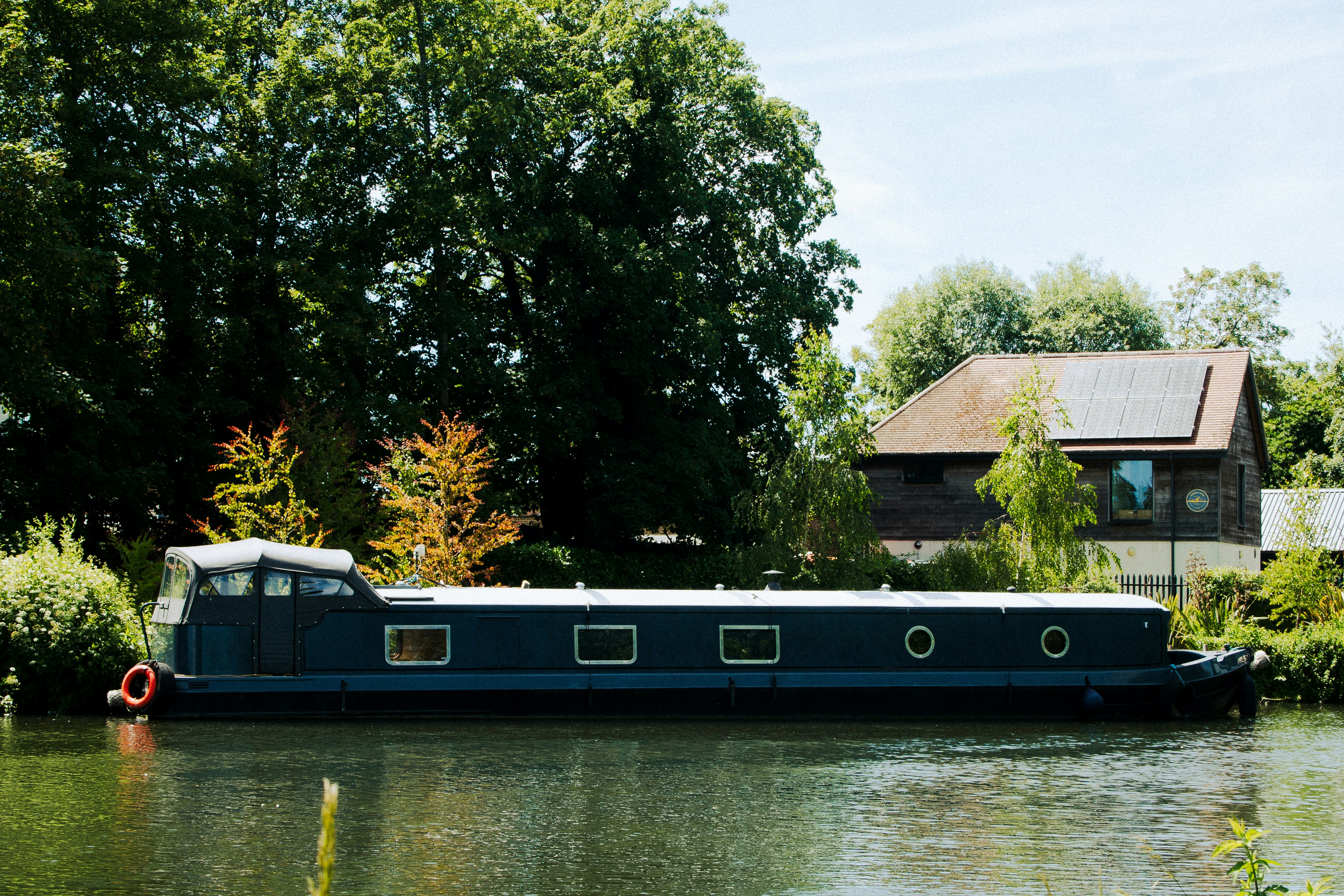 Blue canal narrowboat moored along a calm river with trees and a house in the background.