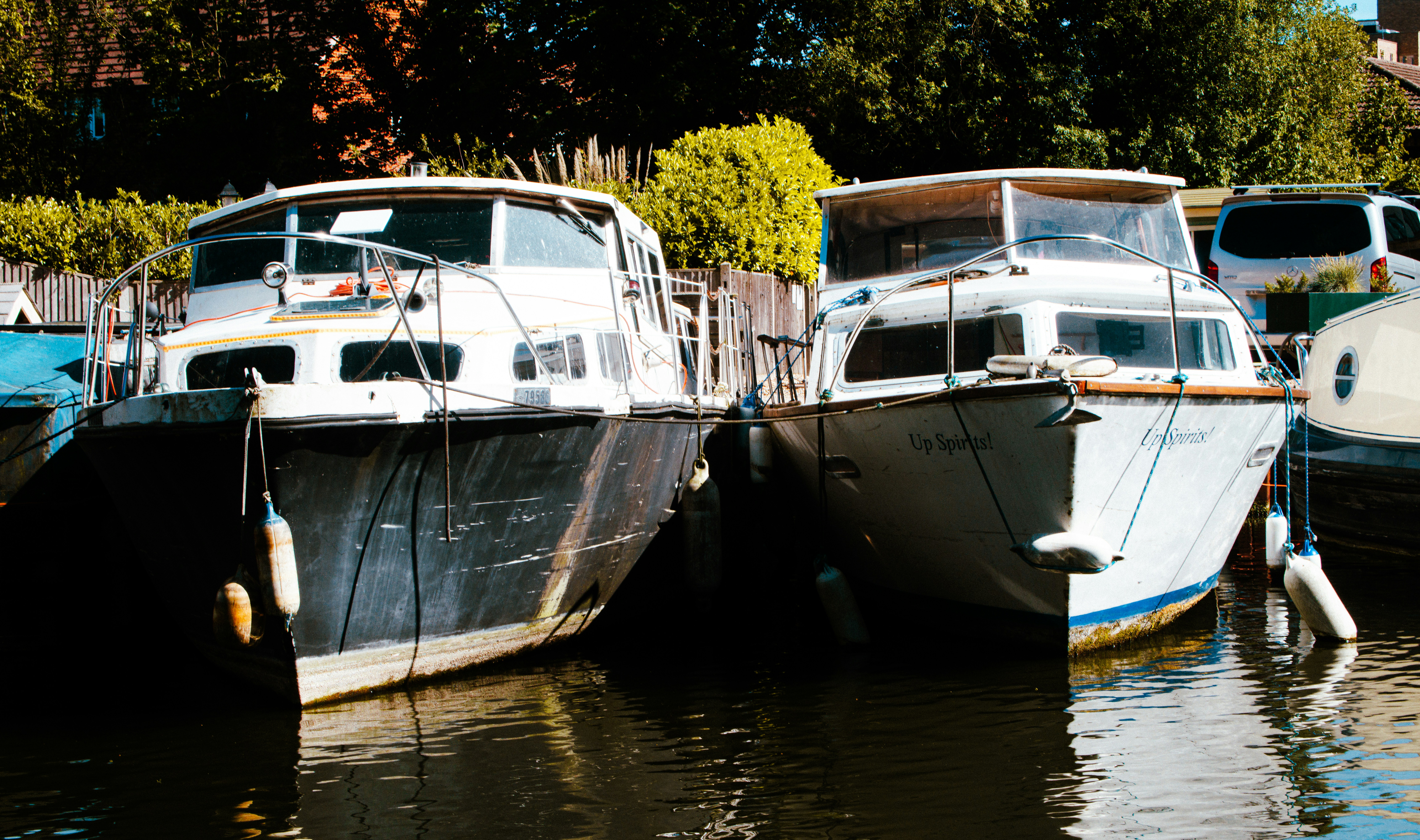 Two motorboats moored side by side at a sunlit marina, their hulls reflecting on calm water.