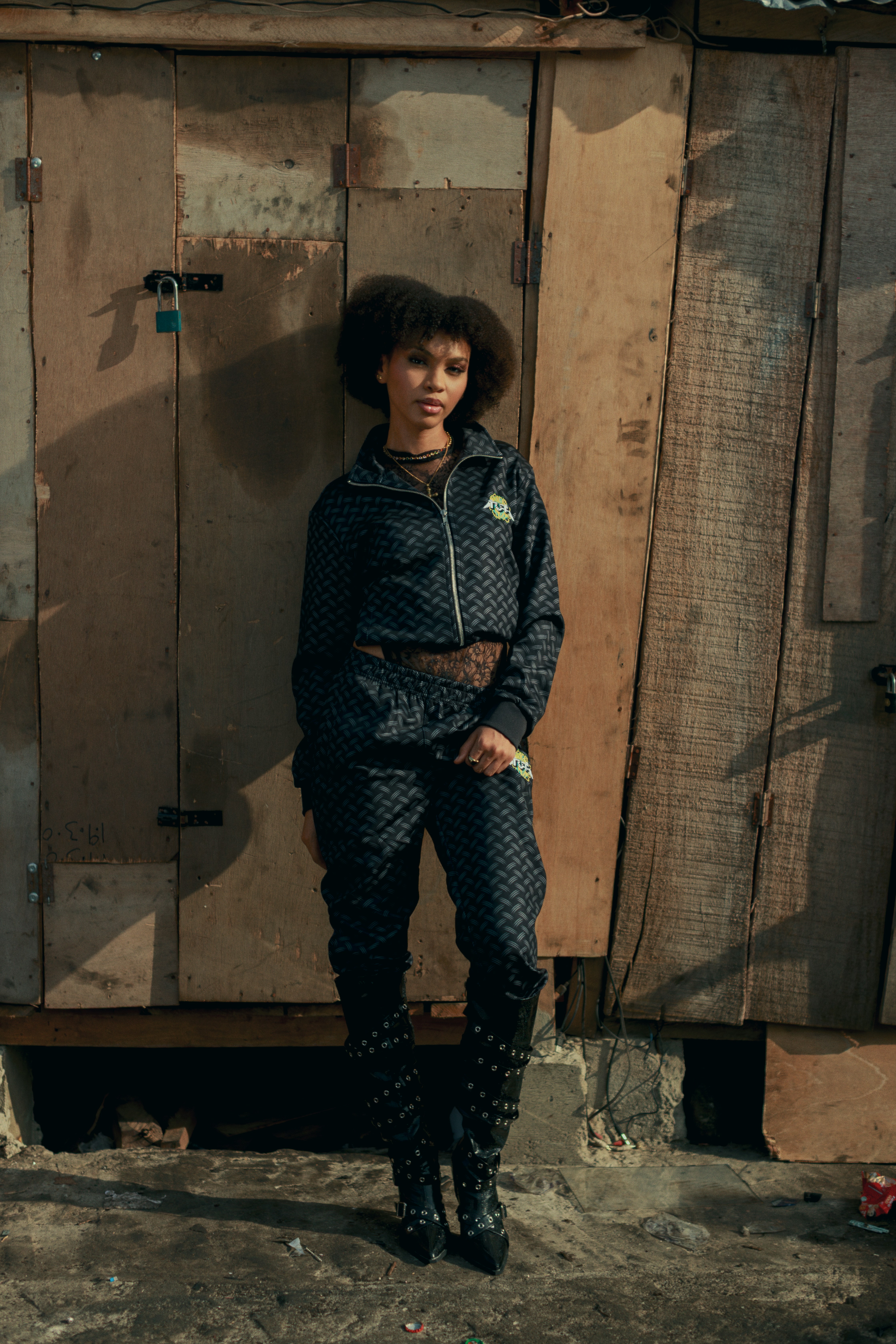 Young woman stands in front of weathered wooden doors, wearing a textured navy tracksuit and tall studded boots; the scene feels gritty yet stylish. A city-edge portrait with a focus on attitude and texture.