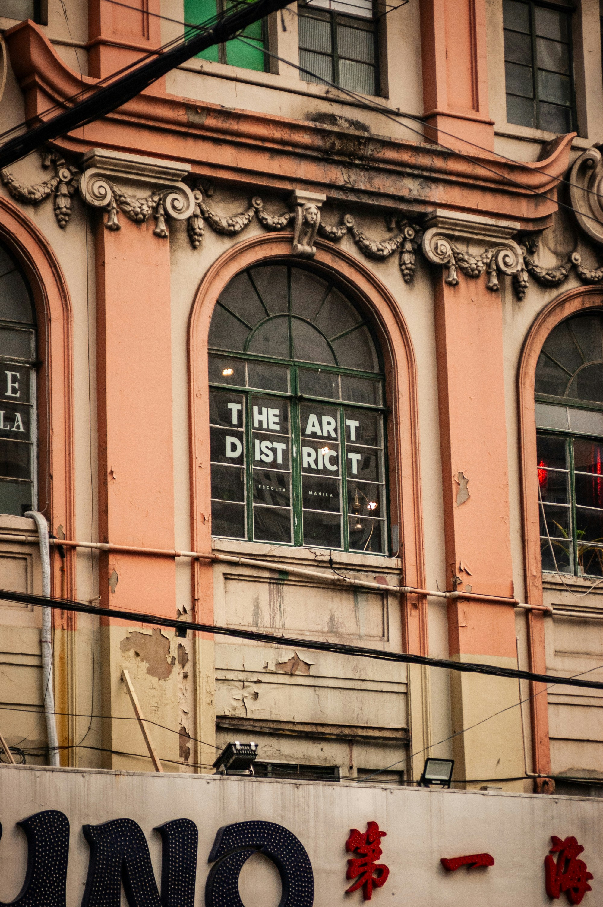 Close-up of an ornate peach-toned building facade with large arched windows and decorative molding, featuring a gallery sign in the window. The scene includes peeling paint and overhead wires, hinting at urban wear. Logo/ storefront signage at the bottom edge.