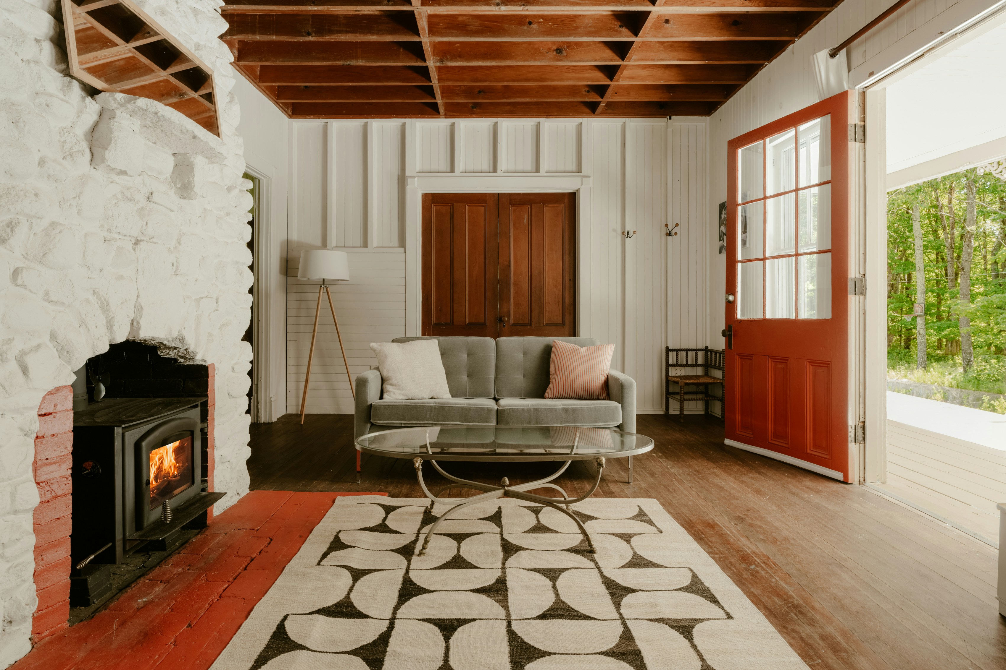 Sunlit living room with a gray sofa, patterned rug, and a wood-burning fireplace; red door open to a lush outdoor view, giving a warm, inviting atmosphere. Photograph showing interior architectural details and natural light. 