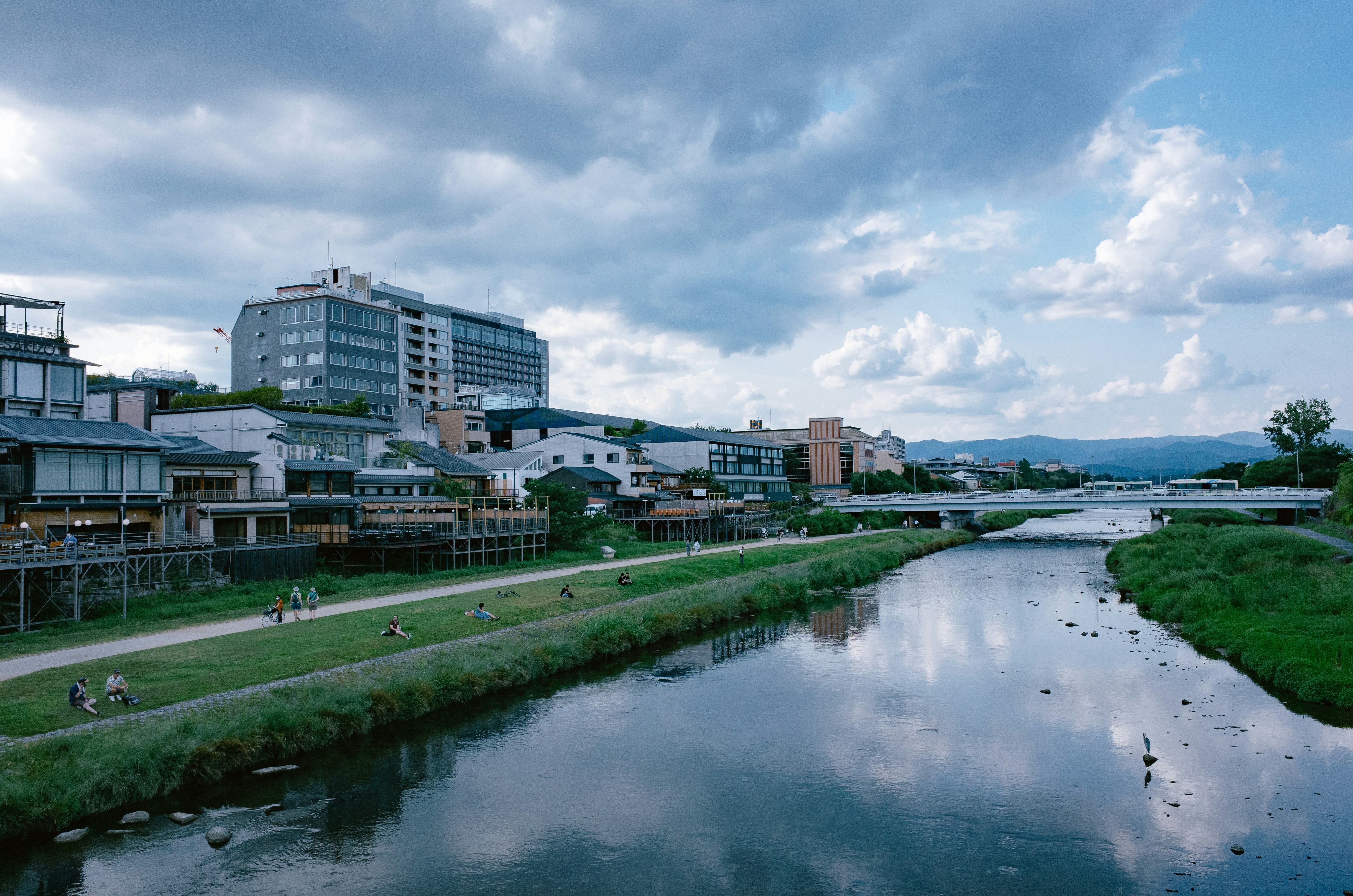 Riverside path along a calm canal with modern buildings on the left and a pedestrian bridge in the distance, under a cloudy sky.