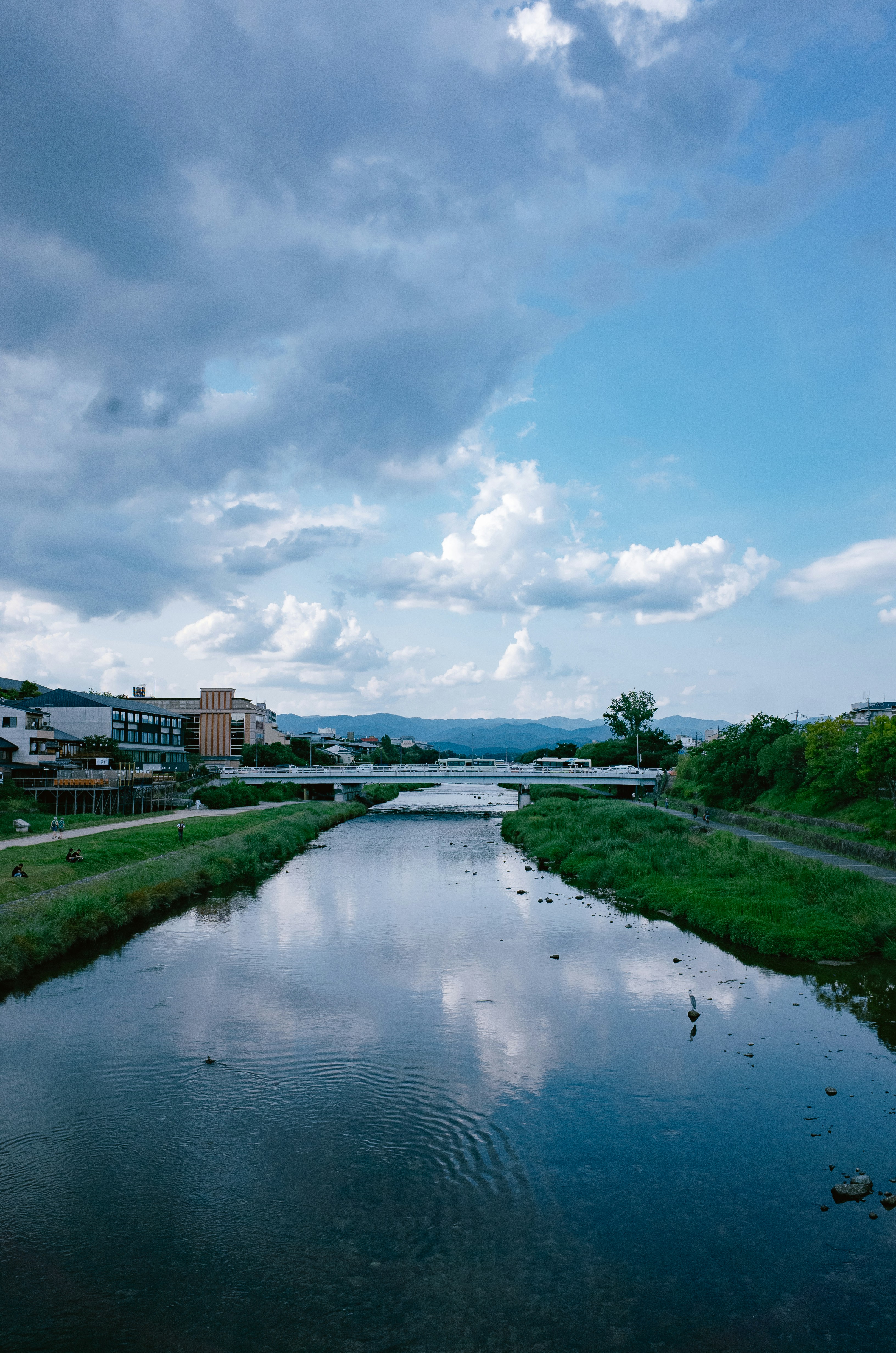 Sunlit river bends through a town with green banks and a distant bridge, reflecting a bright blue sky with clouds.