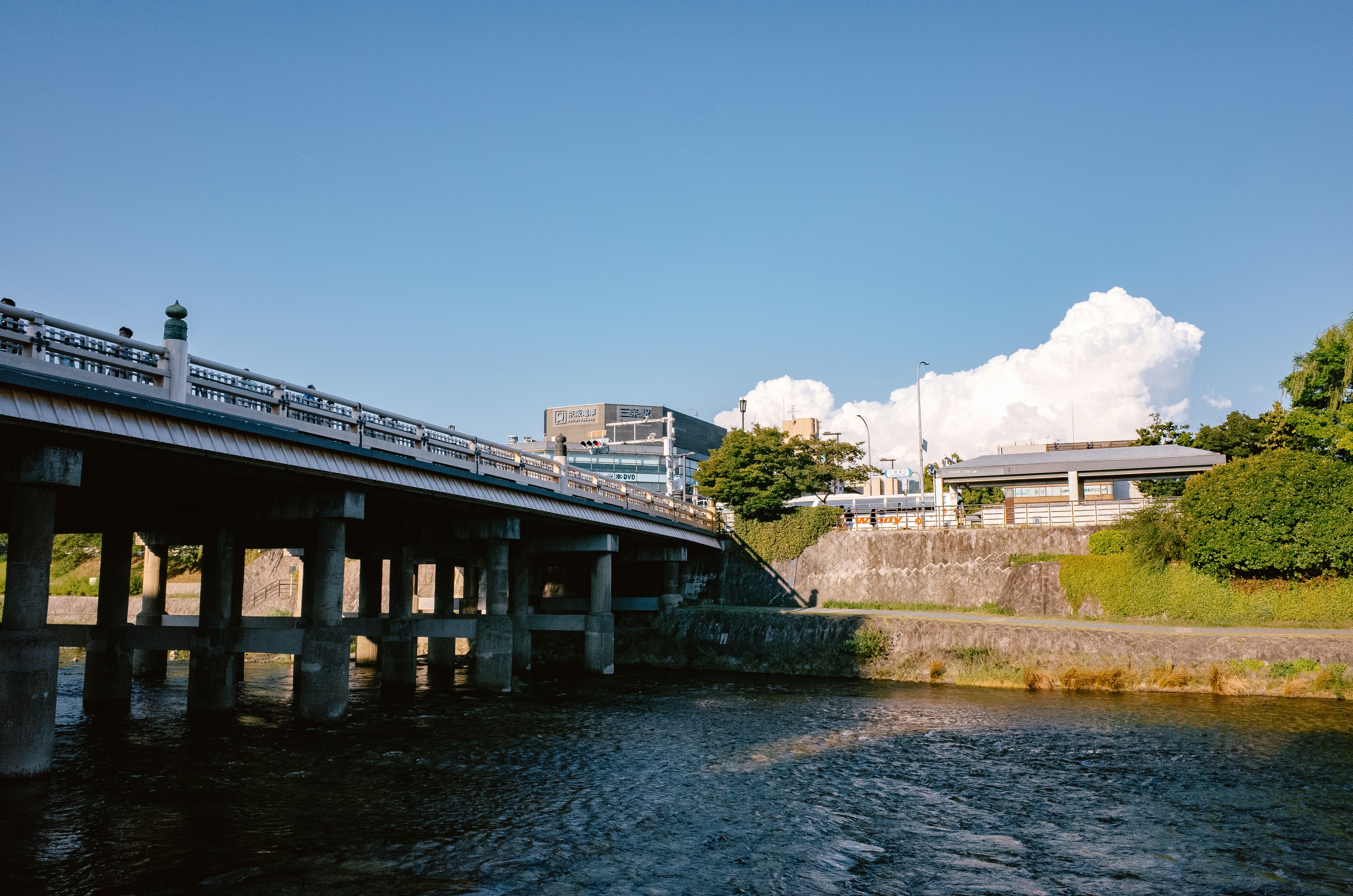 Stone bridge spans a calm river under a clear sky; water lapping against pilings and lush riverside greenery frame the scene.