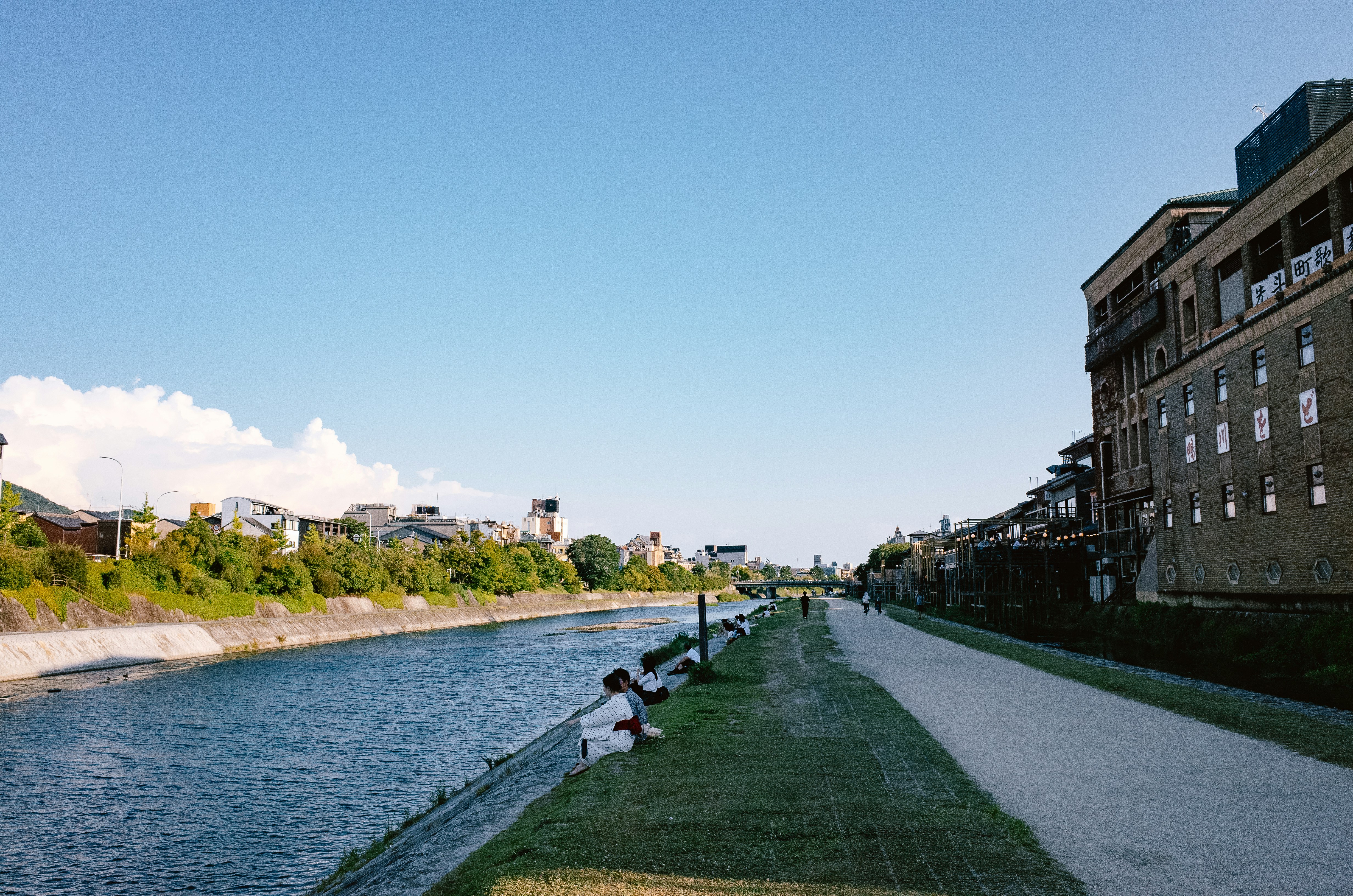 A wide riverfront path beside a calm canal with grassy banks and a row of buildings lining the right side, capturing a serene urban riverside scene.
