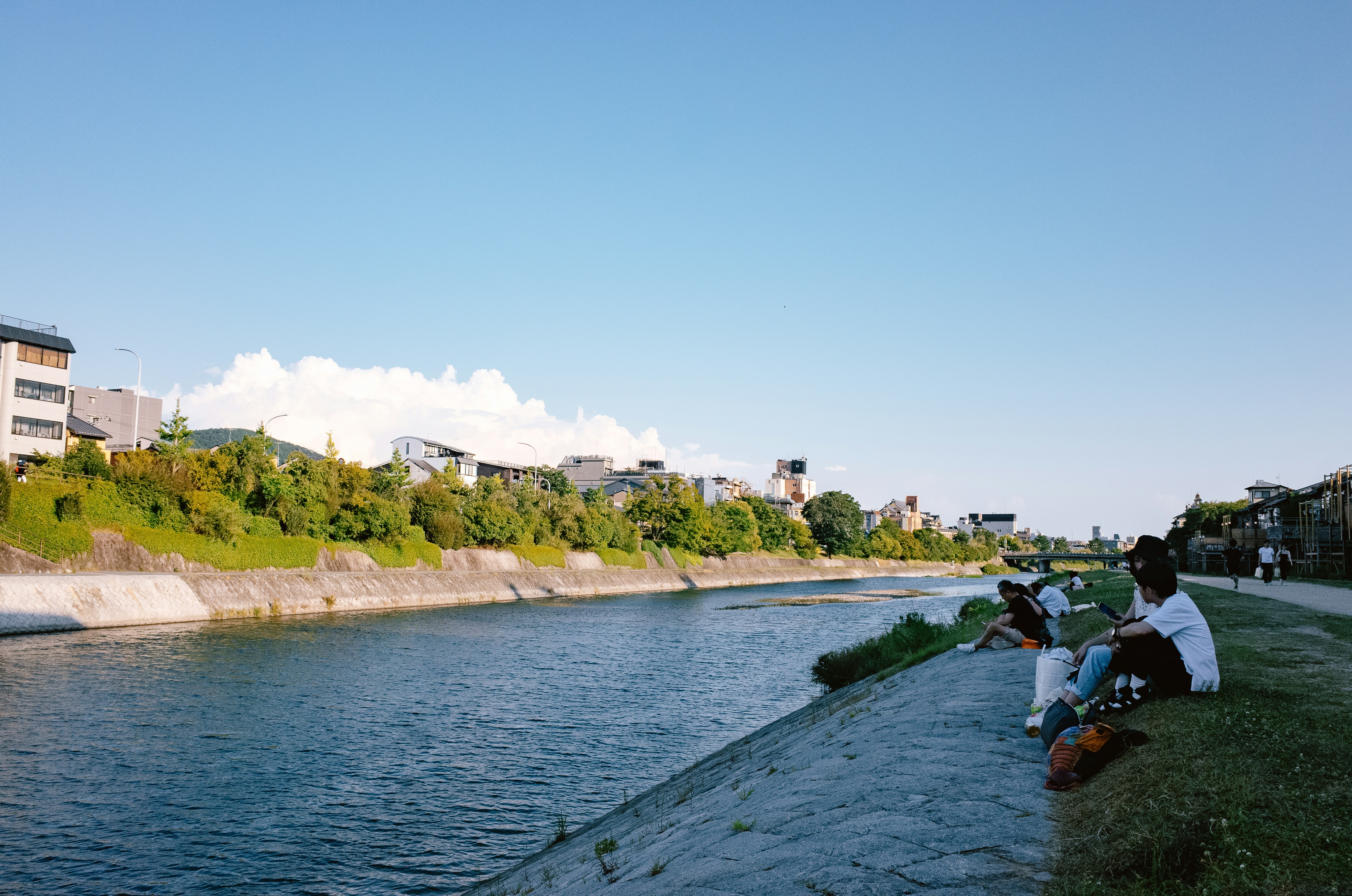 People sit along a riverside embankment, calmly enjoying a sunny day with the cityscape in the distance; a wide-angle scene captures the calm water and grassy verge. This photograph appears as a natural, candid street/landscape shot.