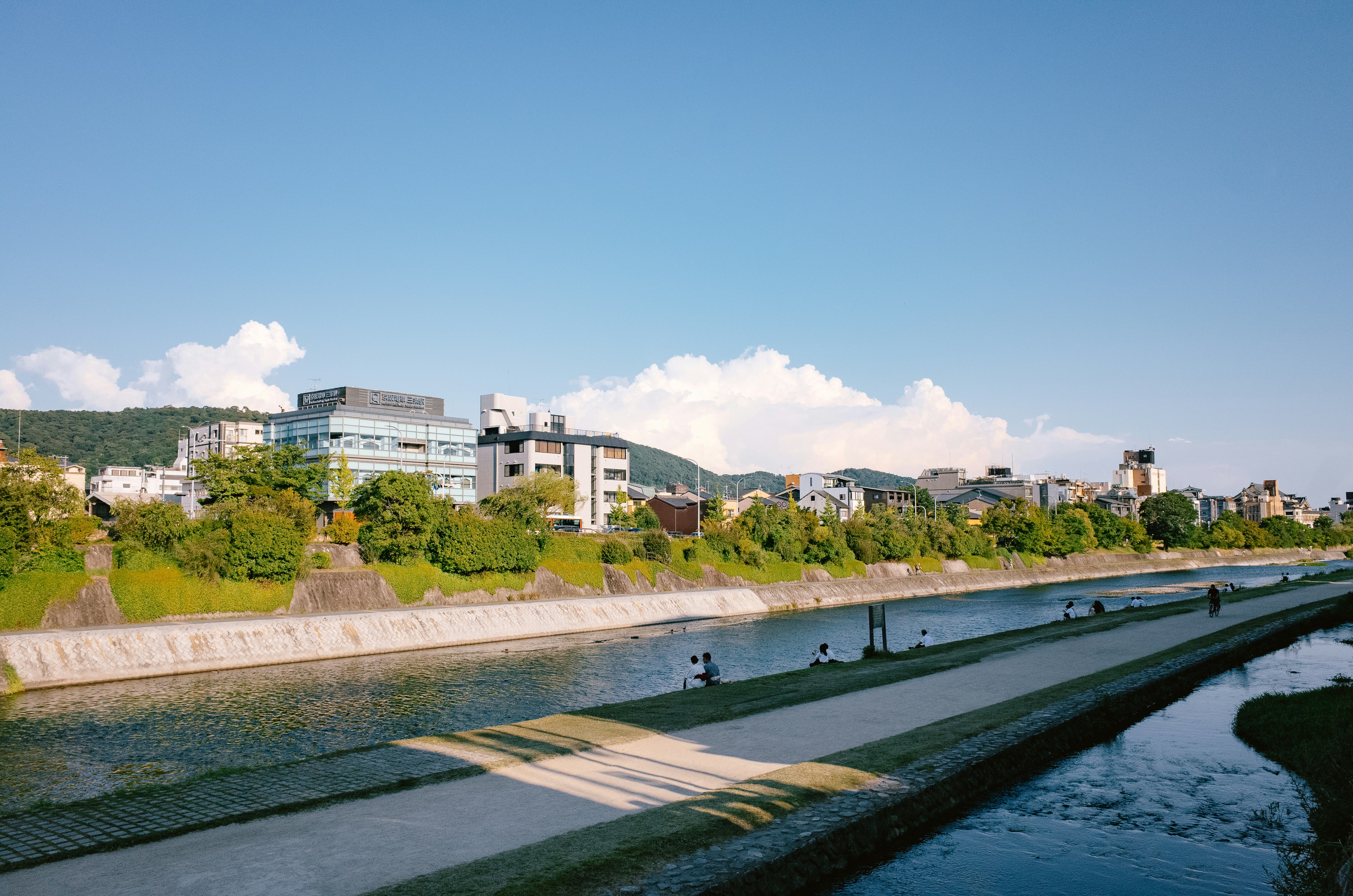 Sunlit riverbank with a paved walkway and modern buildings along the waterline under a blue sky. A calm, urban riverscape image capturing the contrast between natural water and city architecture.