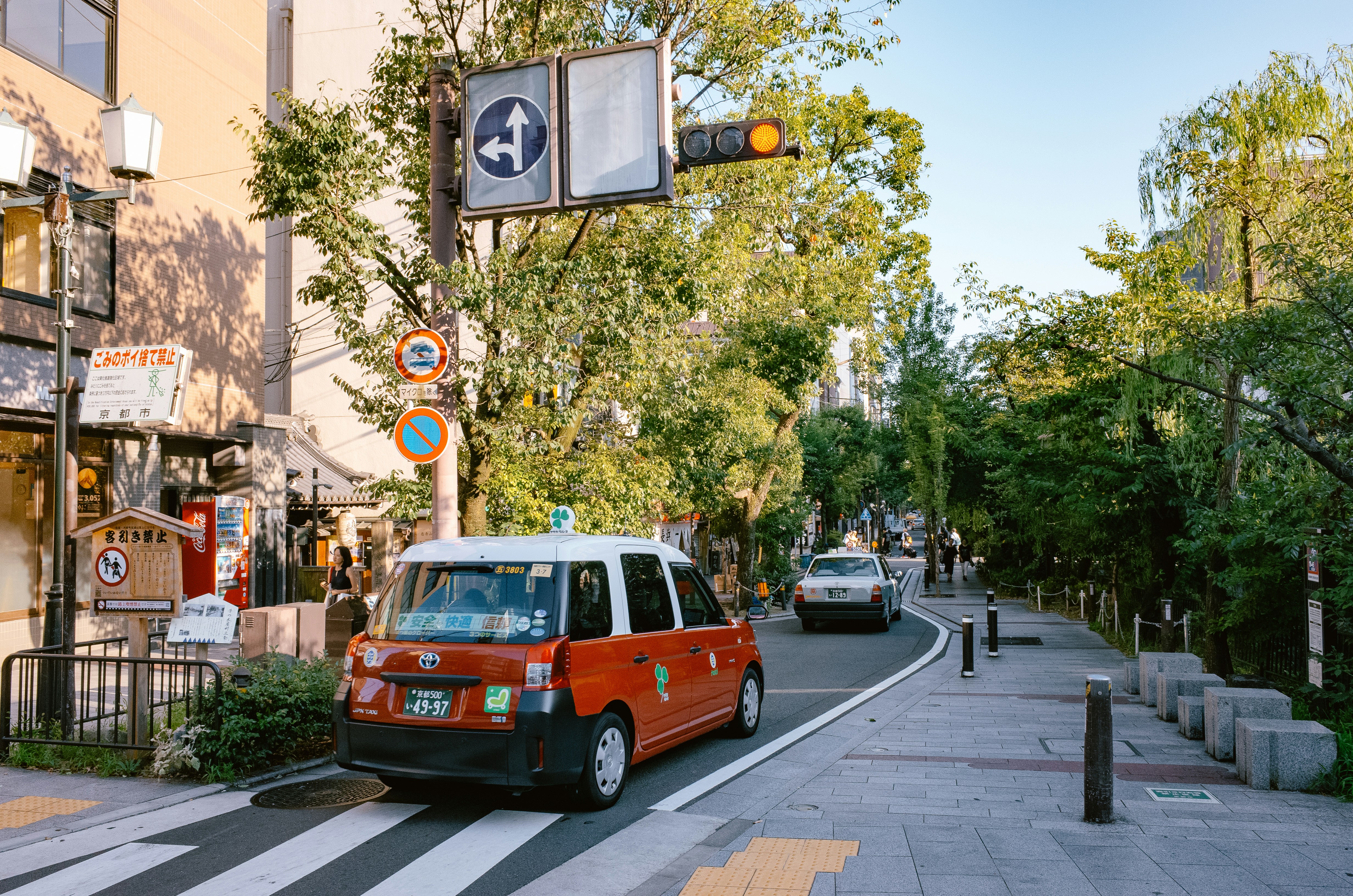Urban street scene featuring an orange van near a pedestrian crosswalk, with storefronts and lush trees on a sunny day.